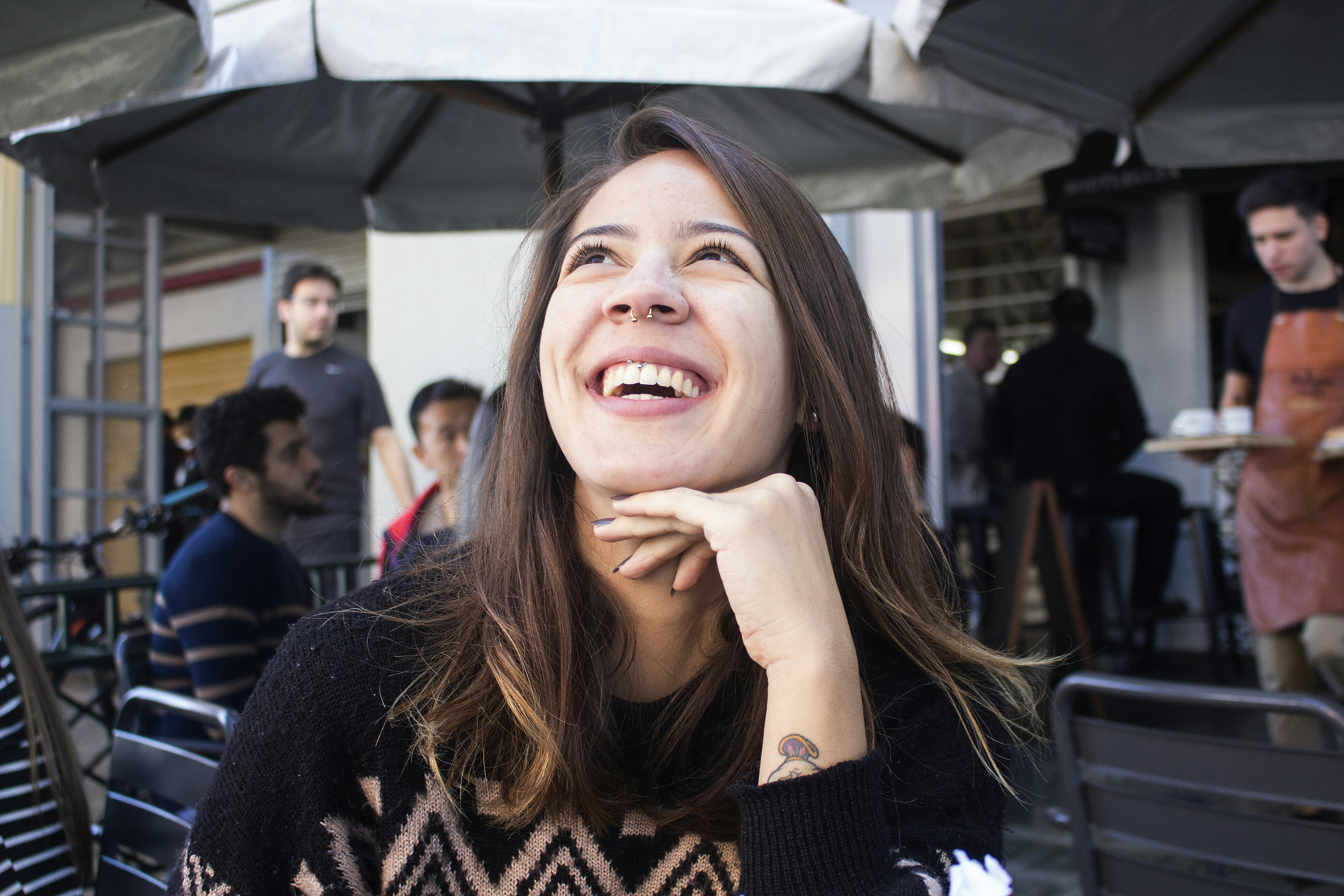 Woman smiling widely while seated outdoors at a cafe under umbrellas.