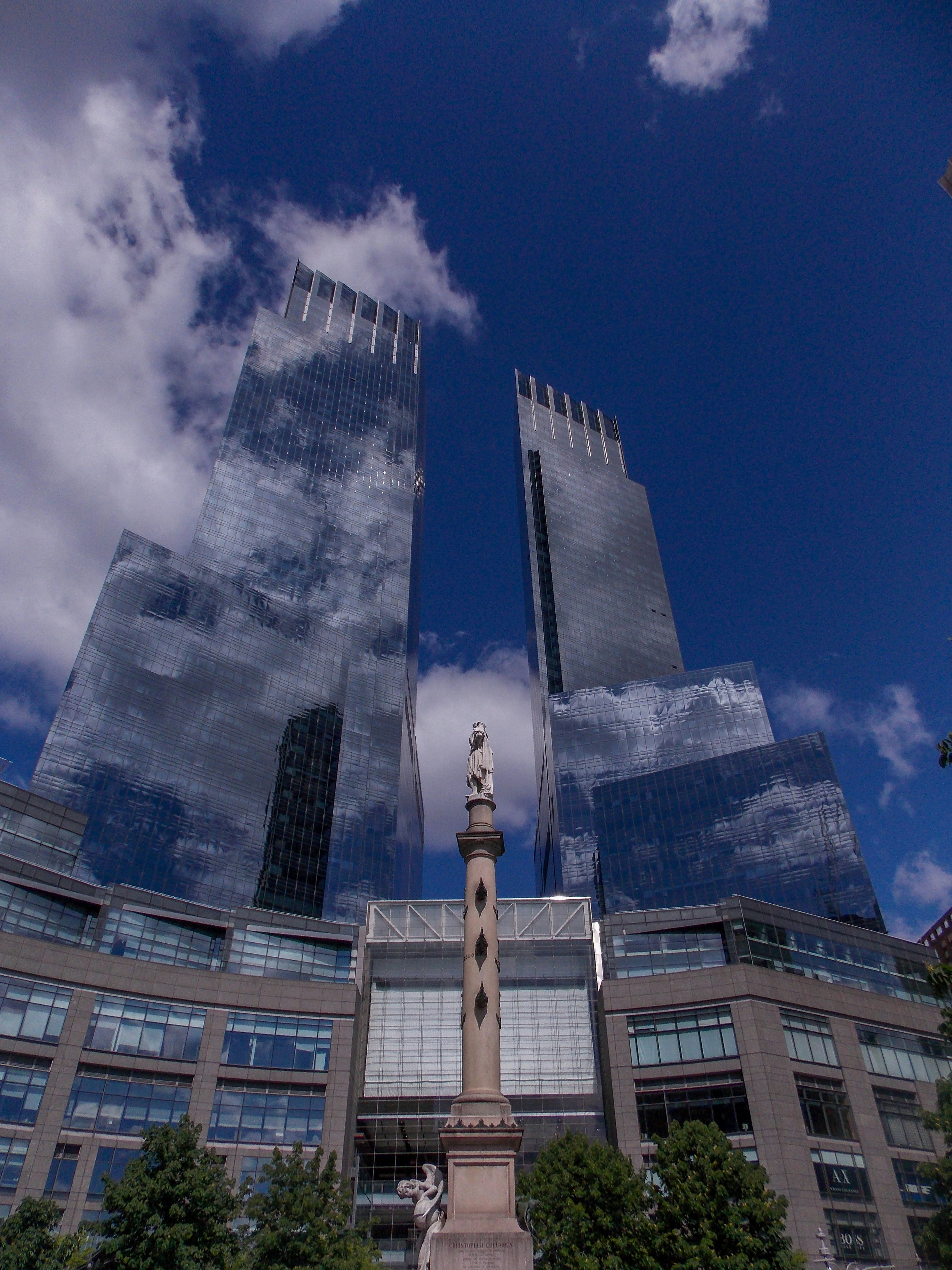A towering monument stands in the foreground while sleek skyscrapers reflect clouds in the background, symbolizing the intersection of history and contemporary architecture.