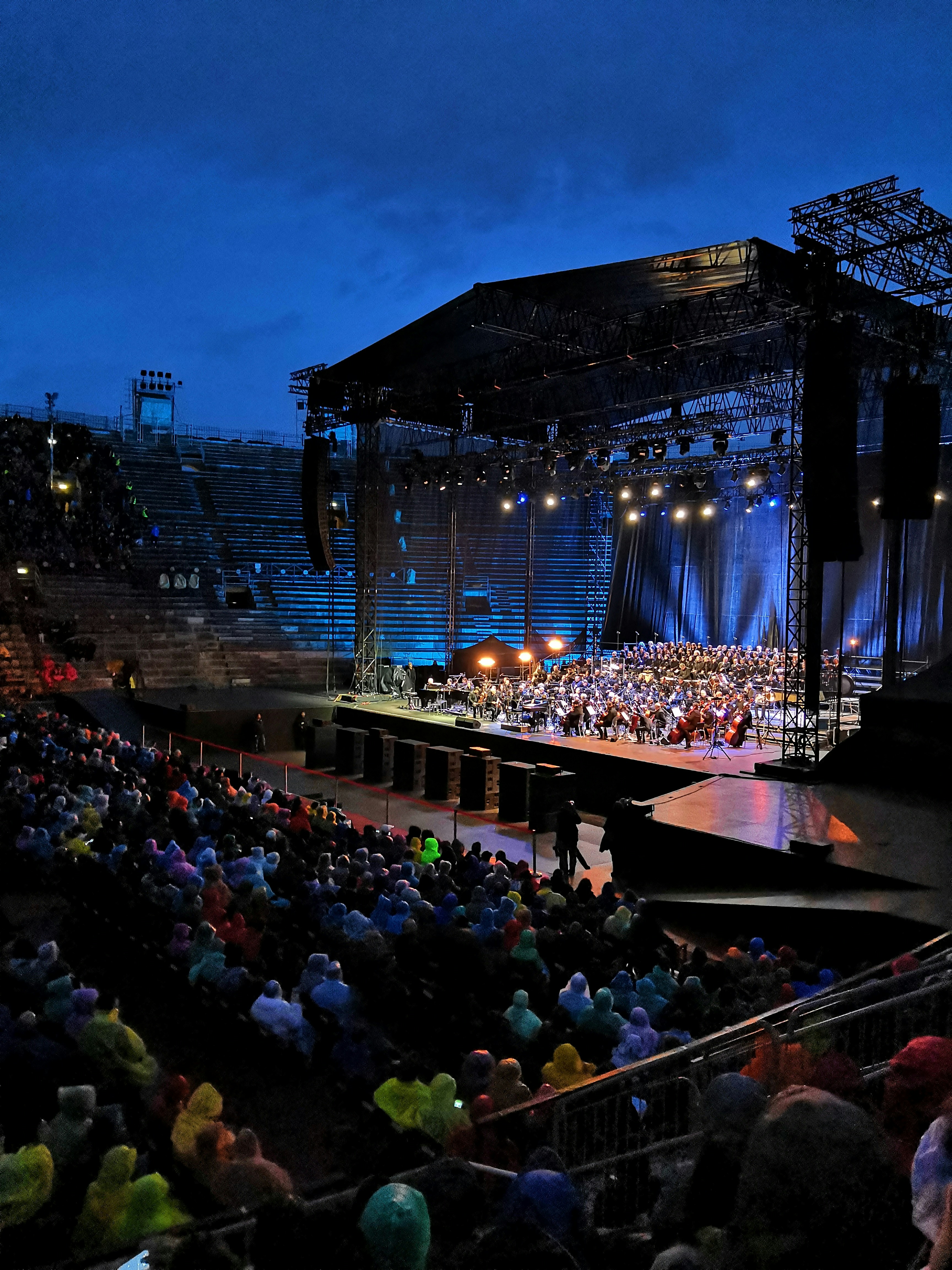 Evening concert at a grand outdoor amphitheater with a vibrant audience in colorful raincoats.