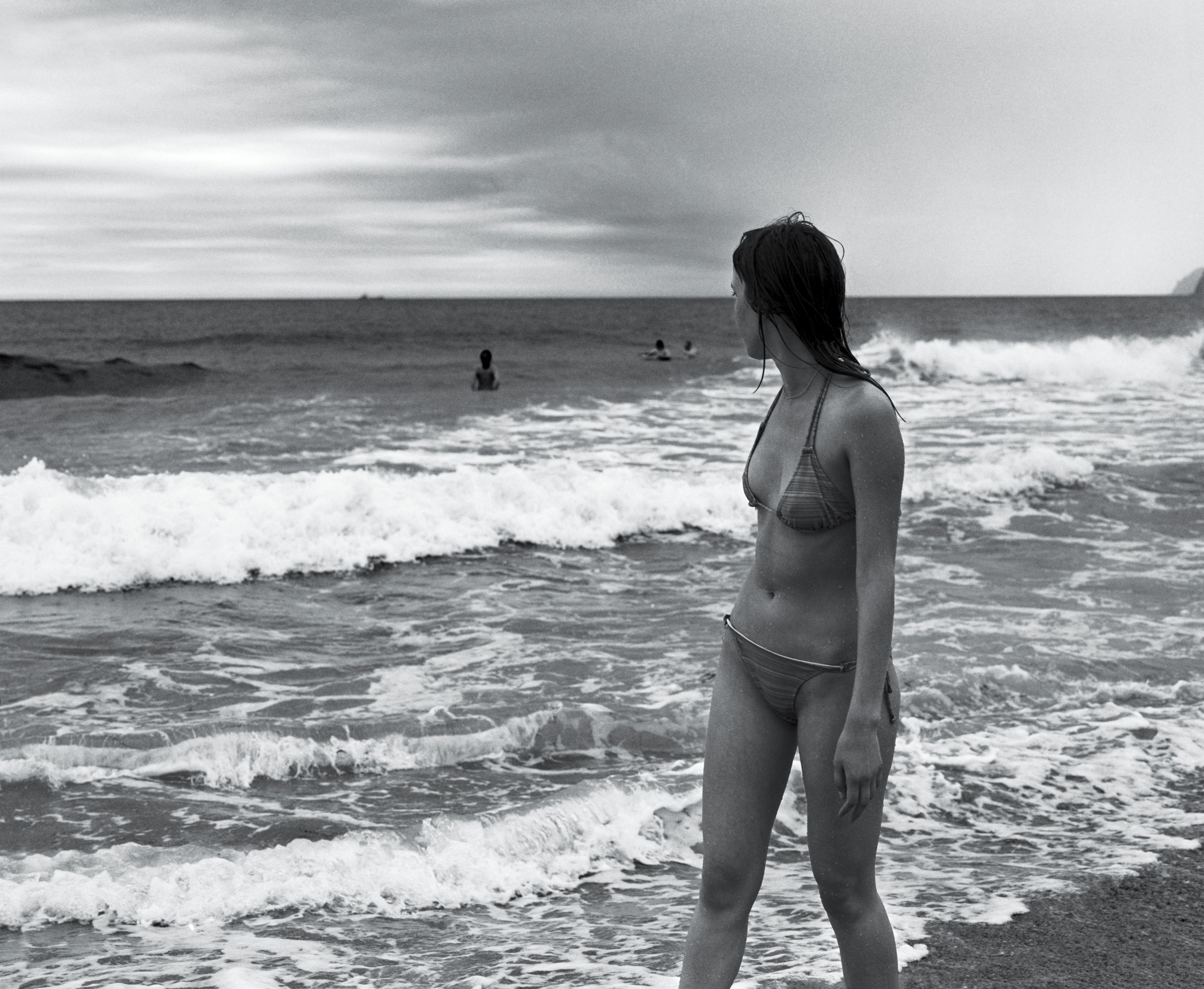 A woman in a bikini stands at the shoreline, gazing at the waves as they crash gently. The scene captures a moment of contemplation by the water's edge.