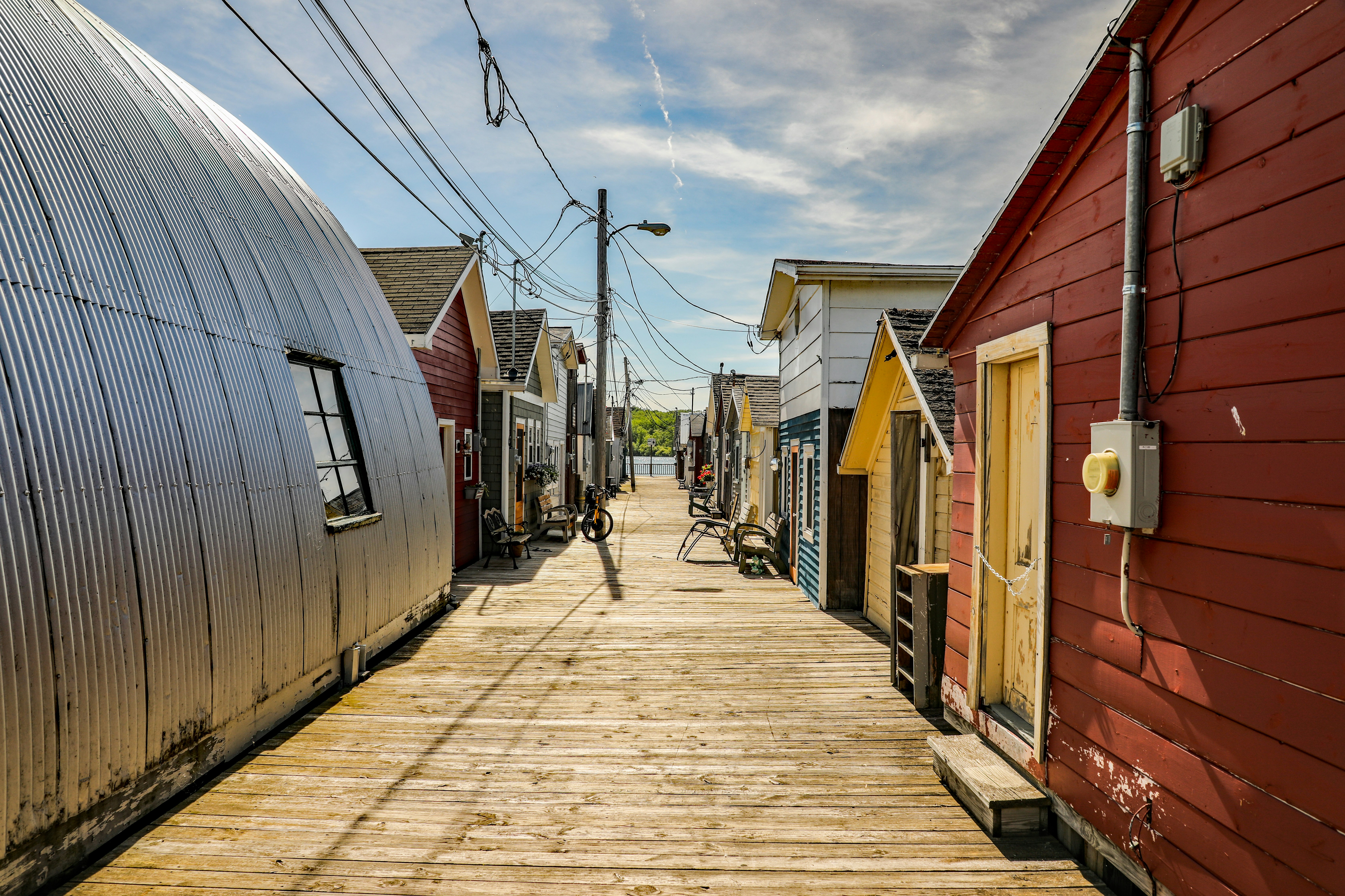 Narrow wooden boardwalk flanked by a curved metal structure and colorful wooden shacks under a partly cloudy sky.