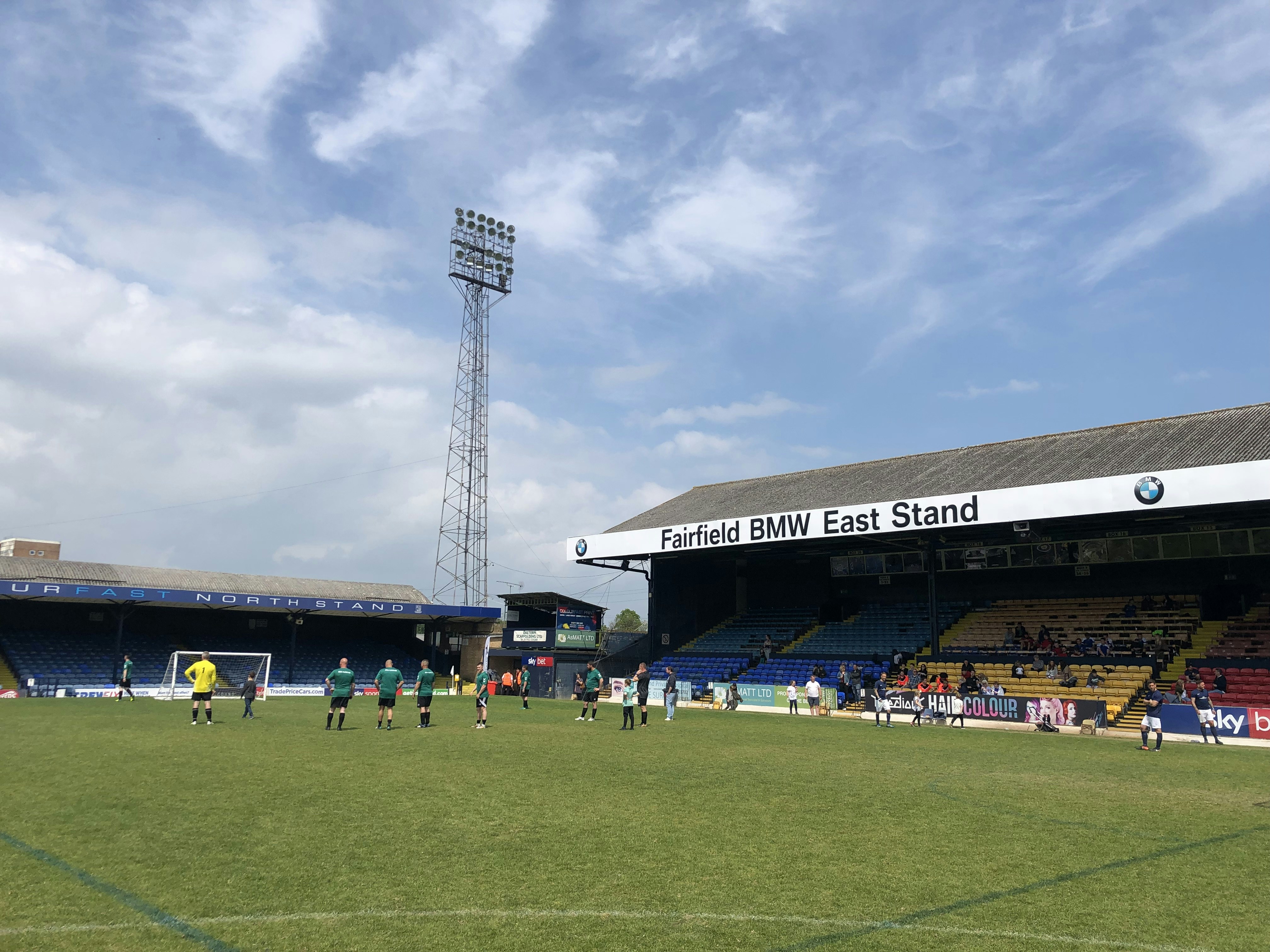 Football players on a grassy field under a partly cloudy sky at Roots Hall Stadium.