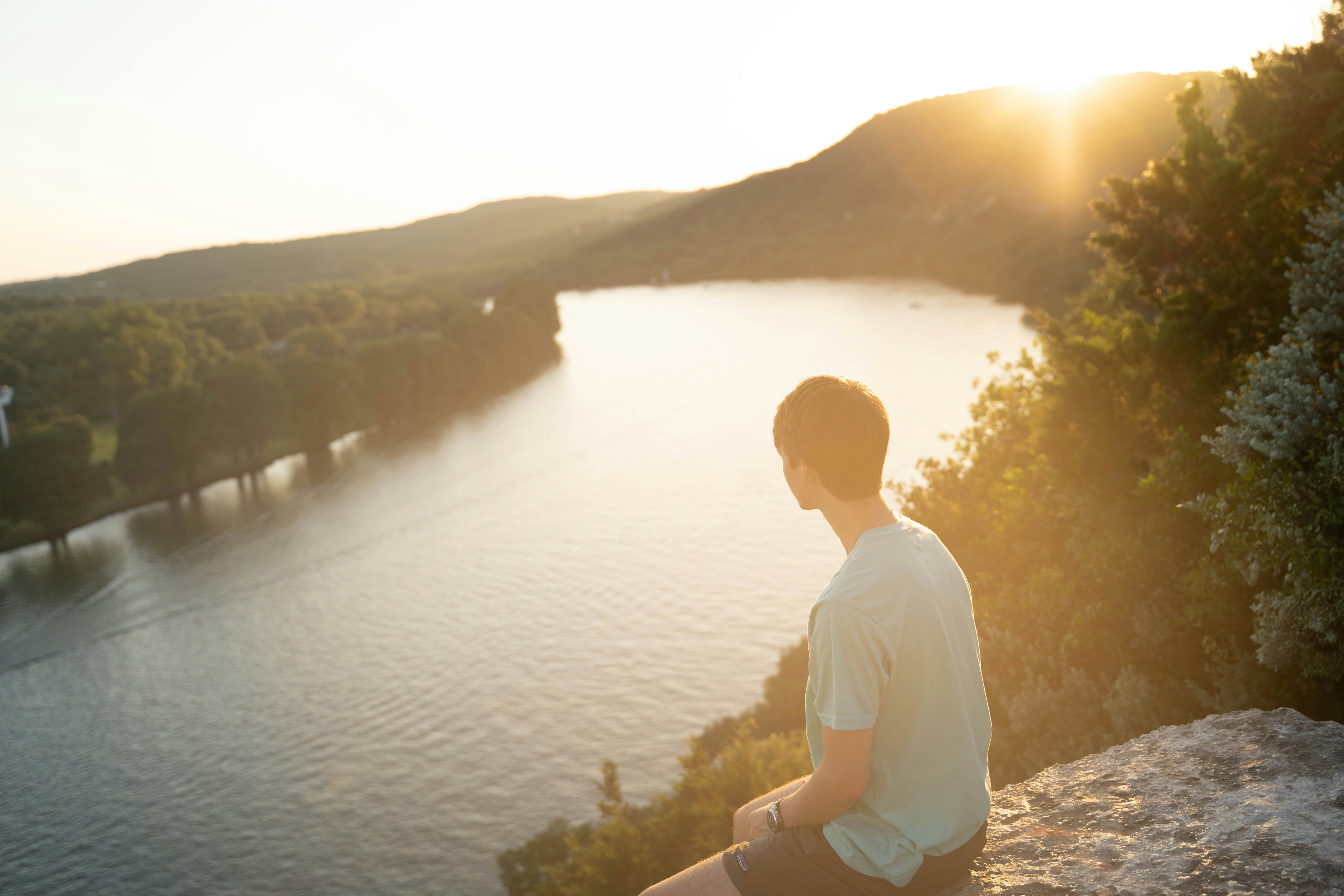 Person seated on a boulder overlooking a sunlit river with distant hills.