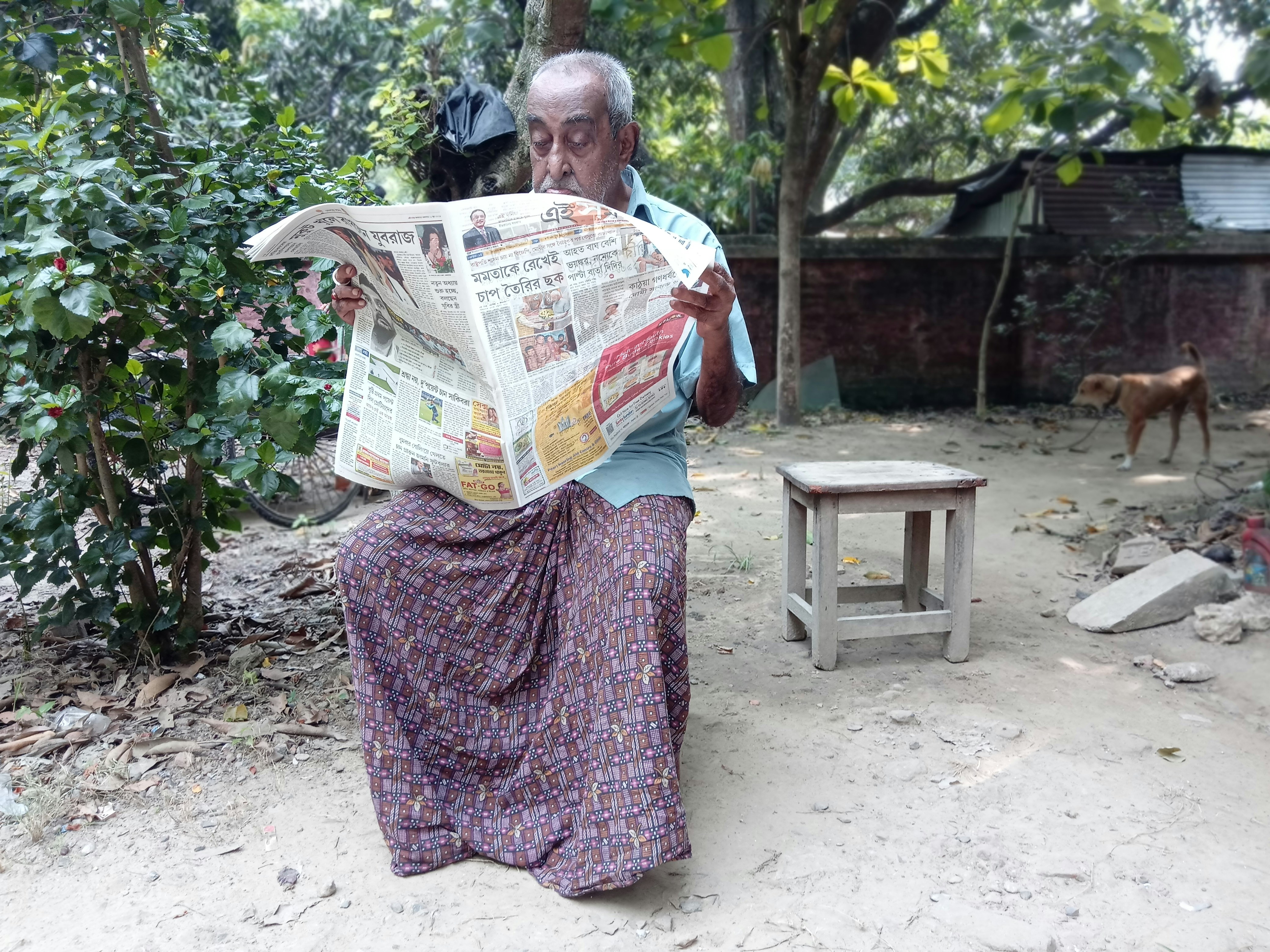 An elderly man sits on the dirt ground reading a newspaper in a sunlit yard, with a dog and lush trees in the background.