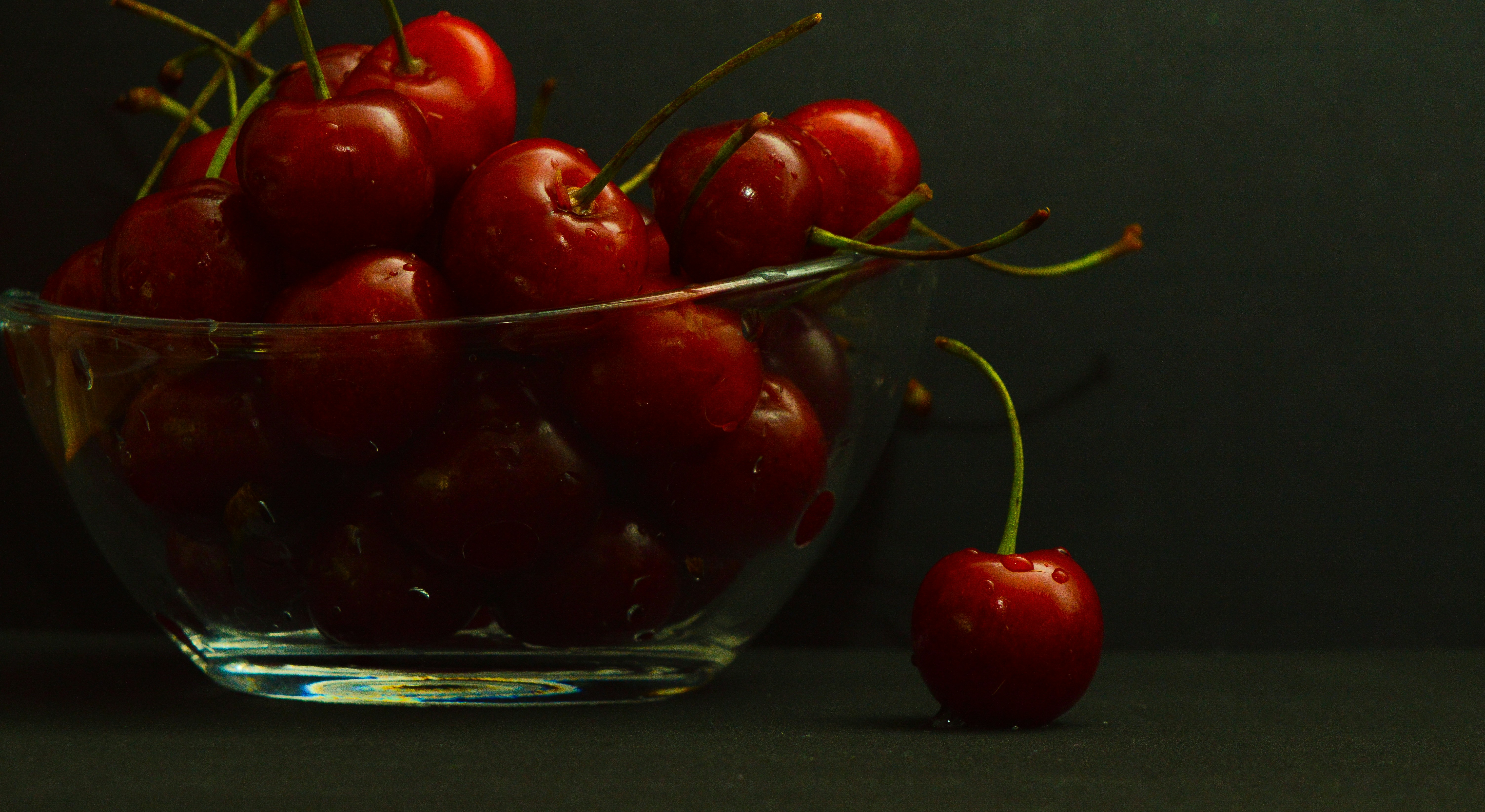 A glass bowl filled with glistening red cherries, with a single cherry resting on a dark surface nearby.