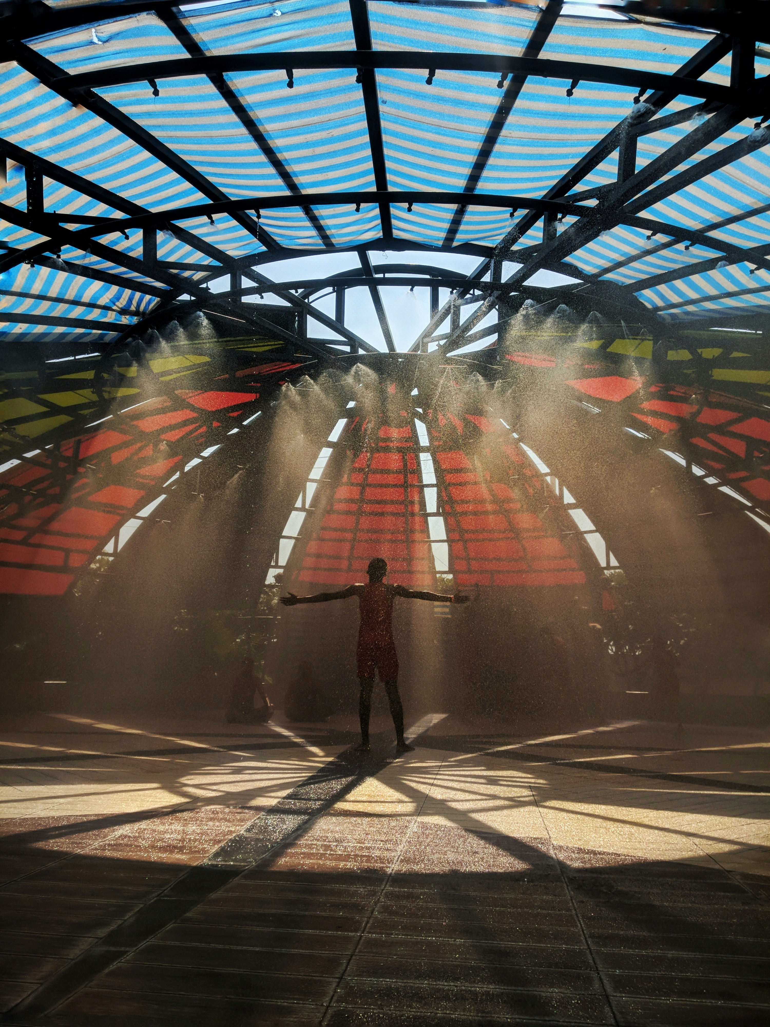 A child stands with arms outstretched in a vibrant water play area, illuminated by beams of sunlight filtering through colorful overhead panels.