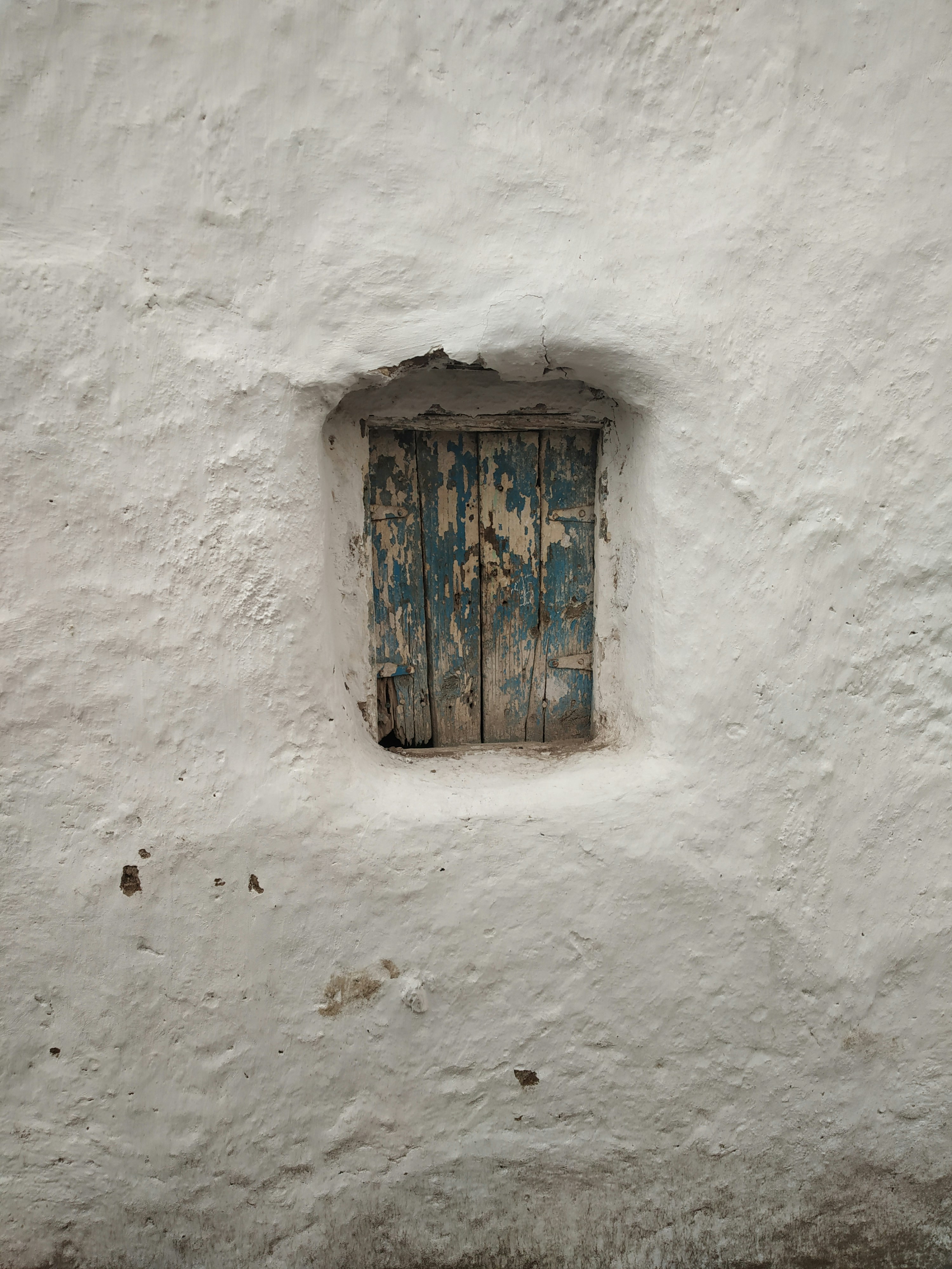 A weathered wooden window framed by a textured white wall, showcasing peeling blue paint and rustic charm.