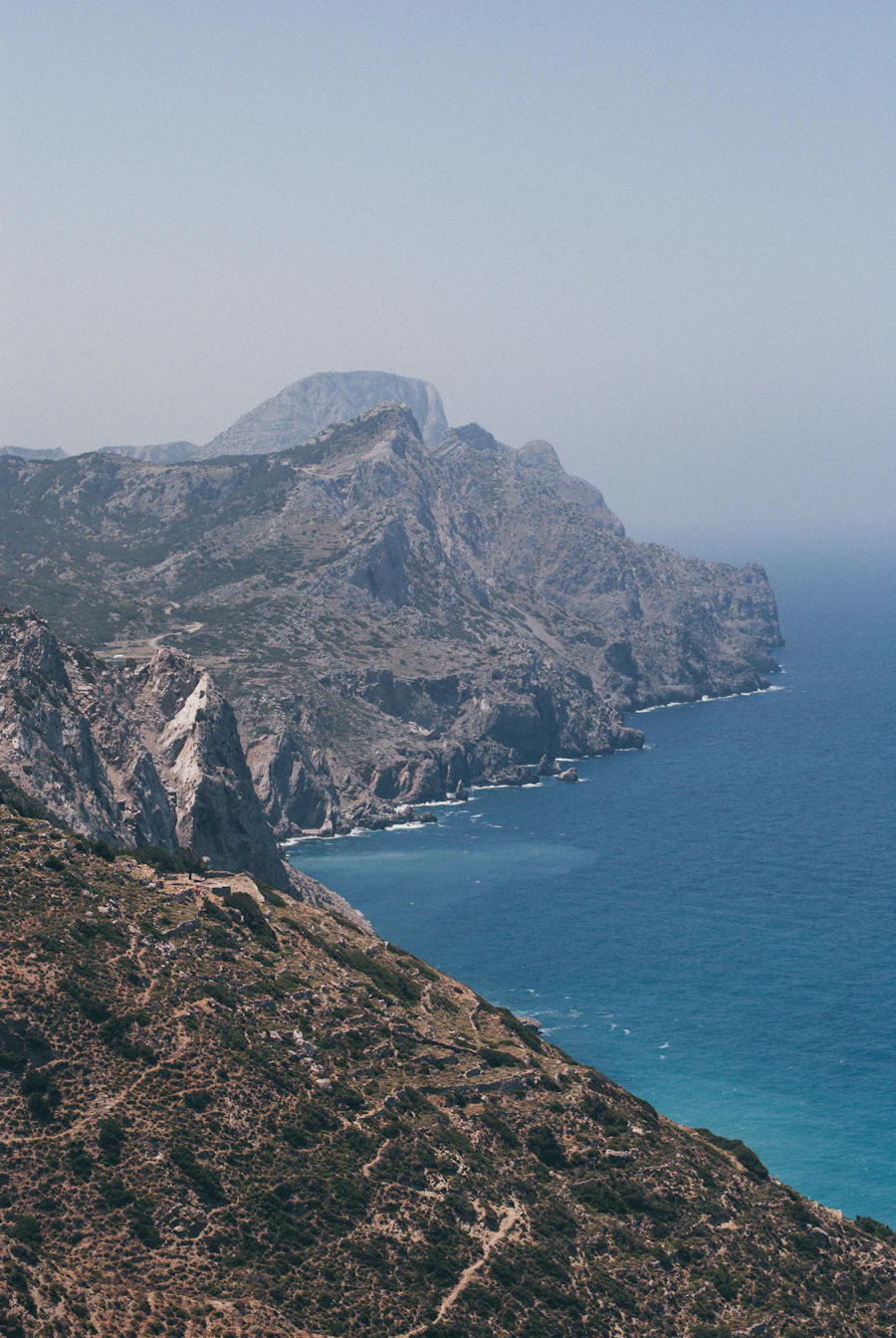 Rugged coastal cliffs jut into a deep blue sea beneath a hazy sky. A steep, scrub-covered foreground ridge leads the eye toward the distant headland.