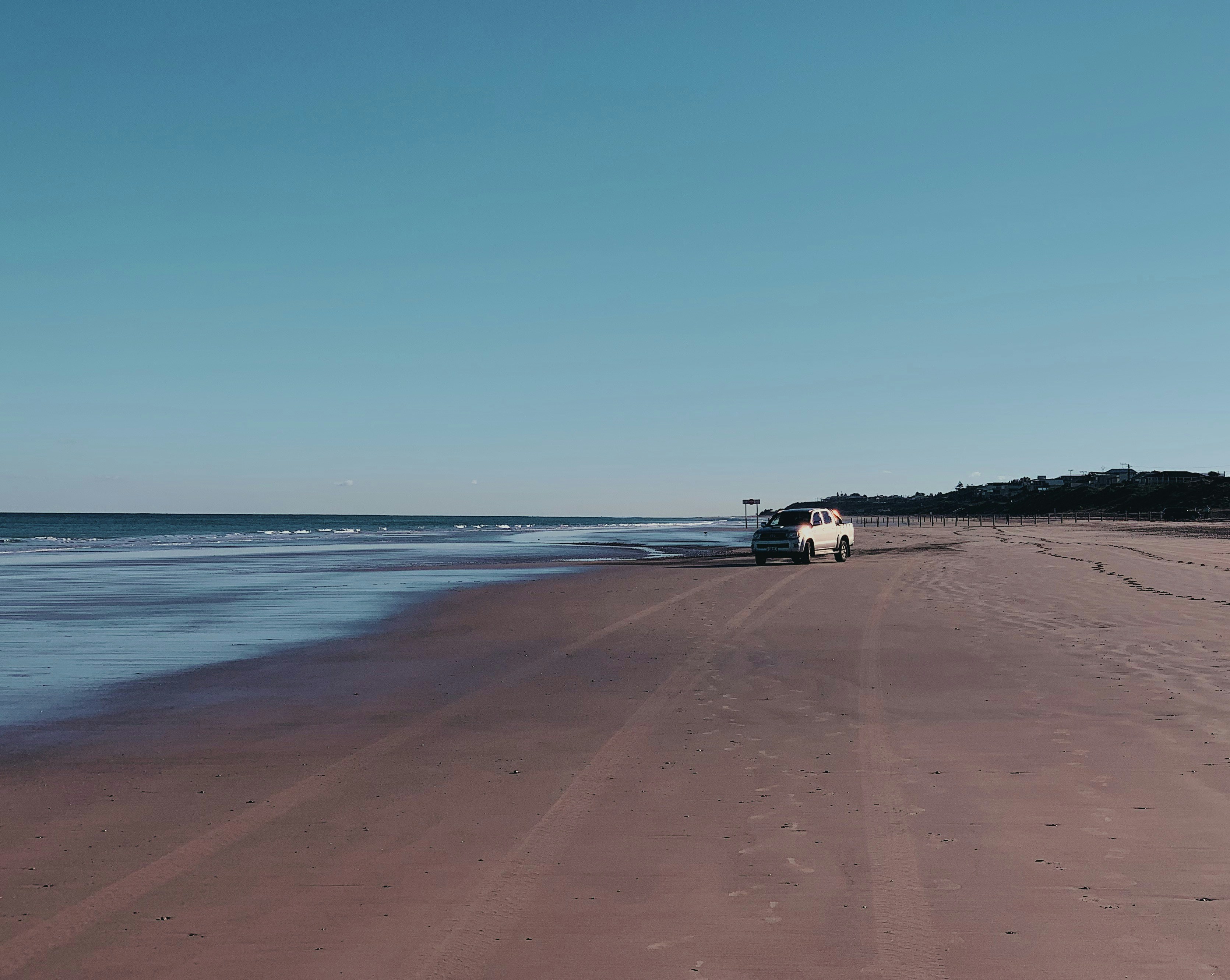 Vehicle traveling along a vast, empty beach under a clear blue sky.