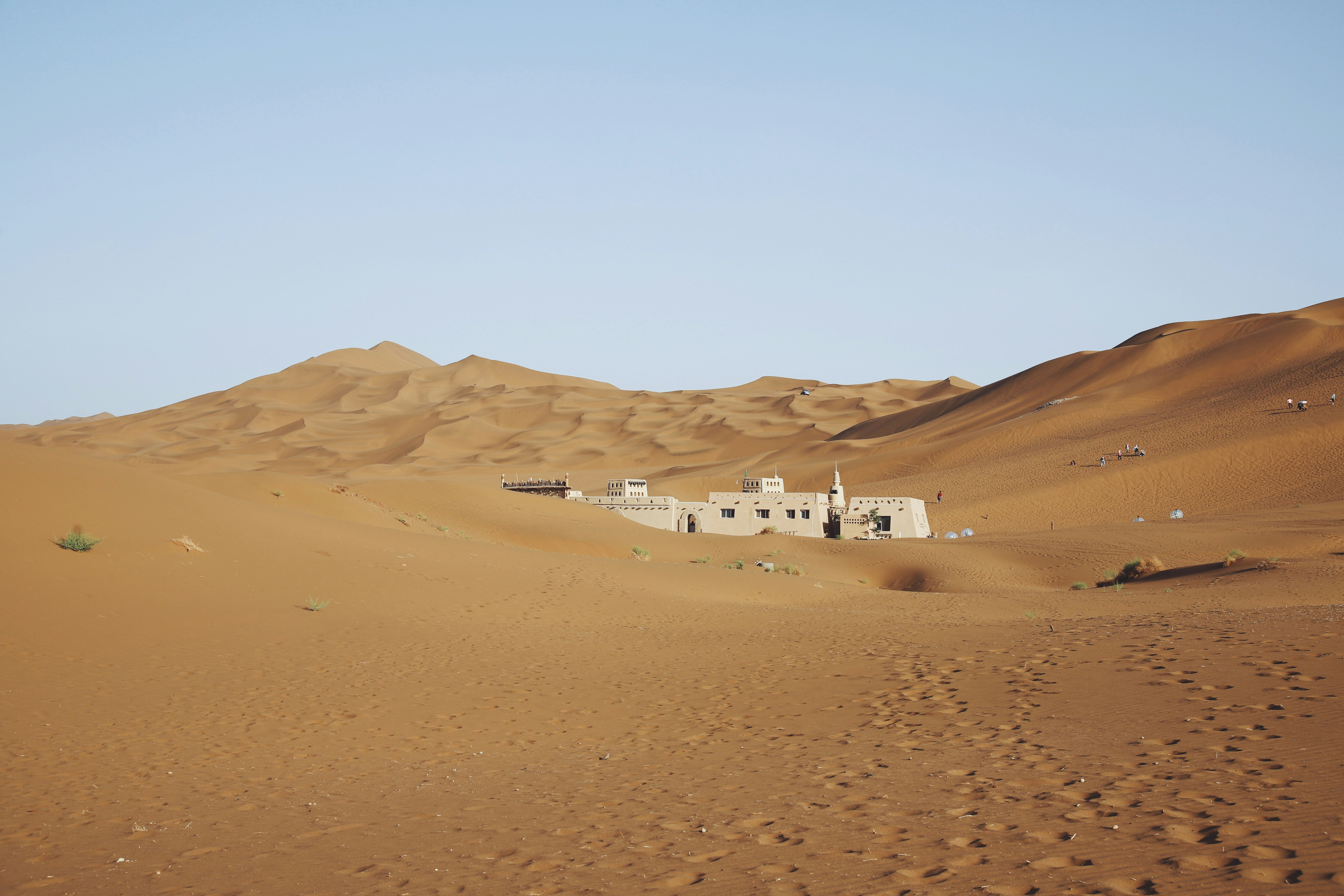 A serene desert landscape featuring a distant structure surrounded by rolling sand dunes under a clear sky.