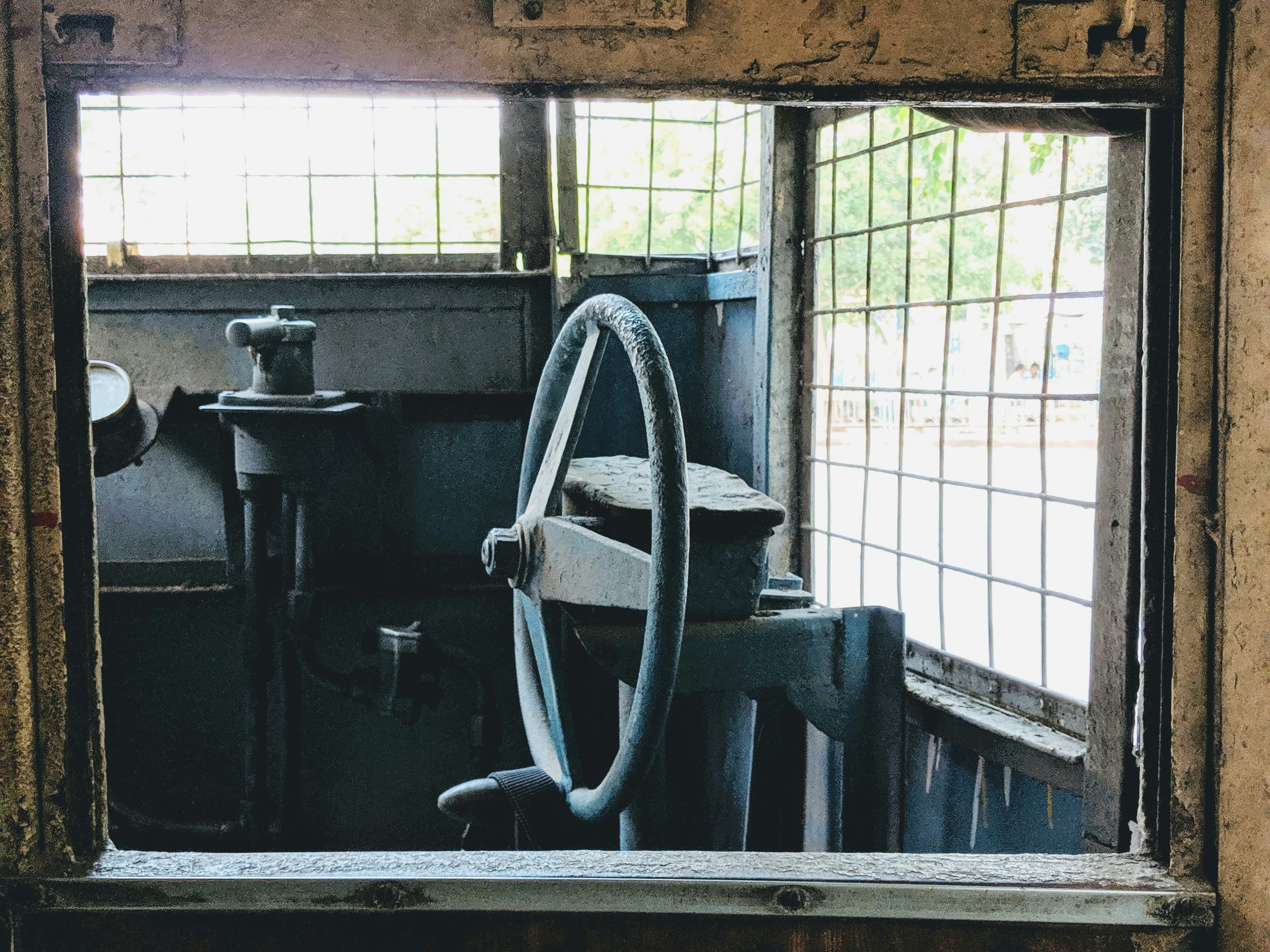Old industrial steering wheel viewed through a rusted, grid-lined window.