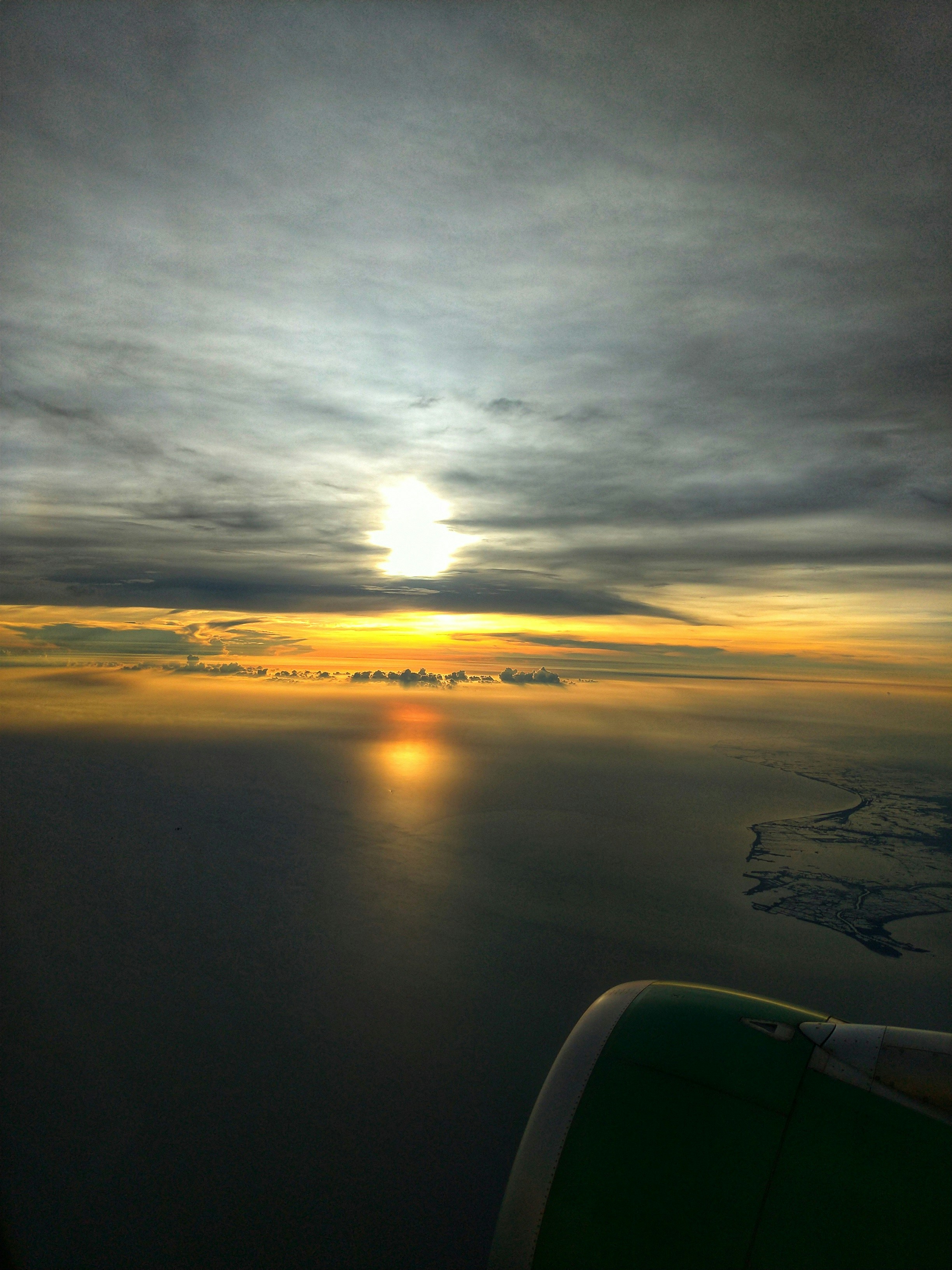 Golden sunlight reflecting on calm ocean waters beneath a blanket of clouds. The airplane wing hints at the journey above the serene landscape.