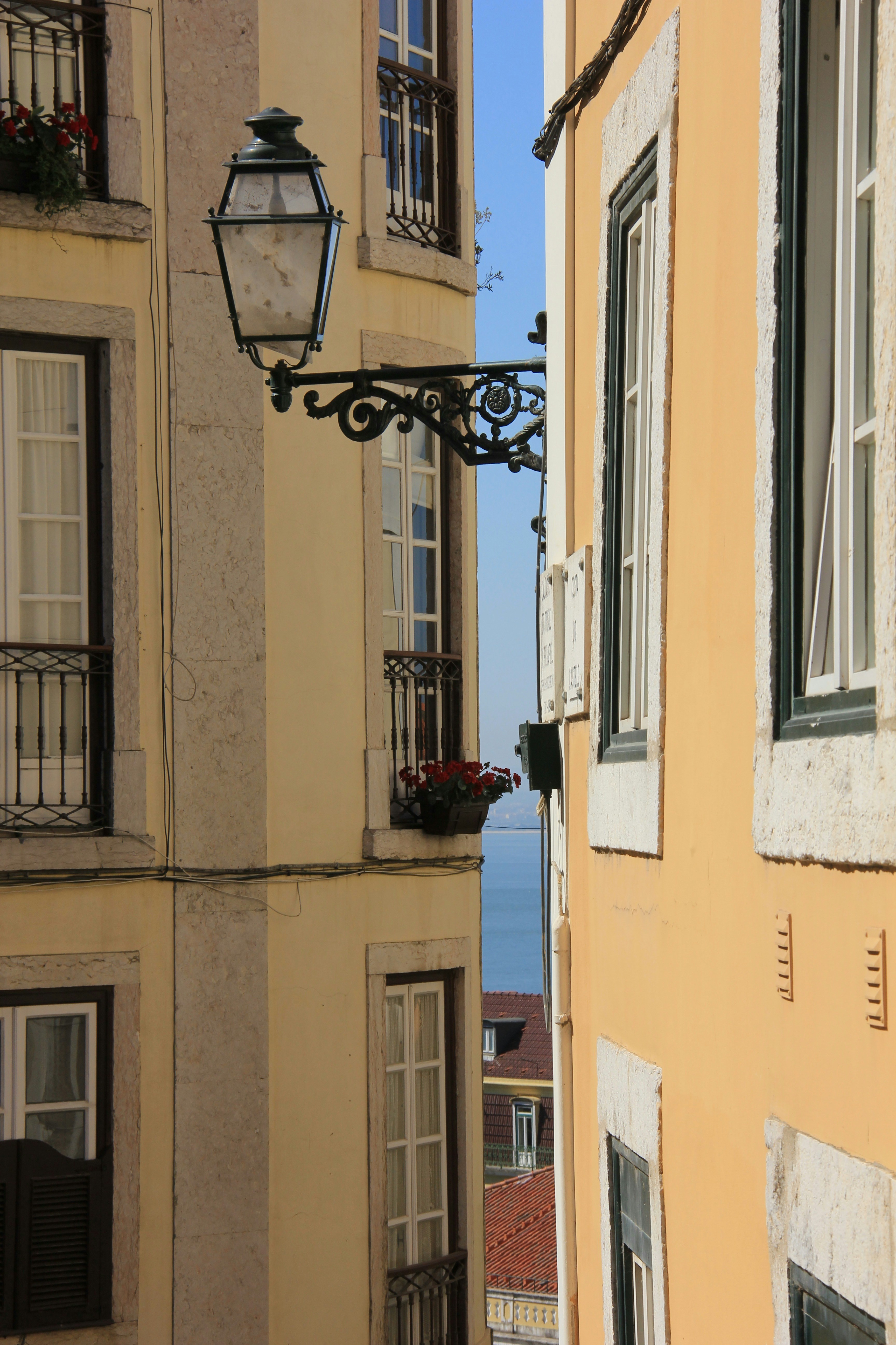 Charming narrow alleyway framed by historical buildings, featuring a vintage lantern and flower-adorned balconies. The scene hints at a coastal view in the distance.