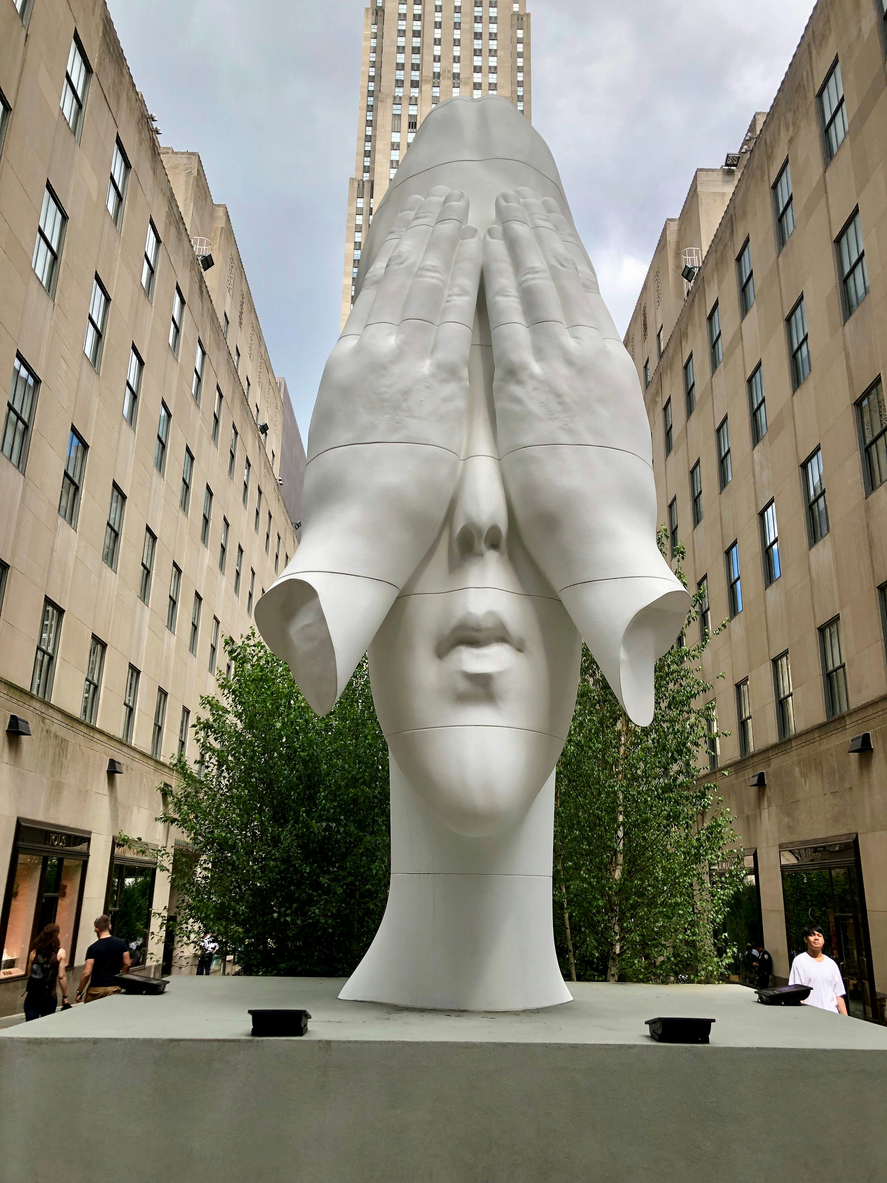 A large white sculpture of a face with hands covering its eyes, set amidst a courtyard surrounded by buildings and greenery.