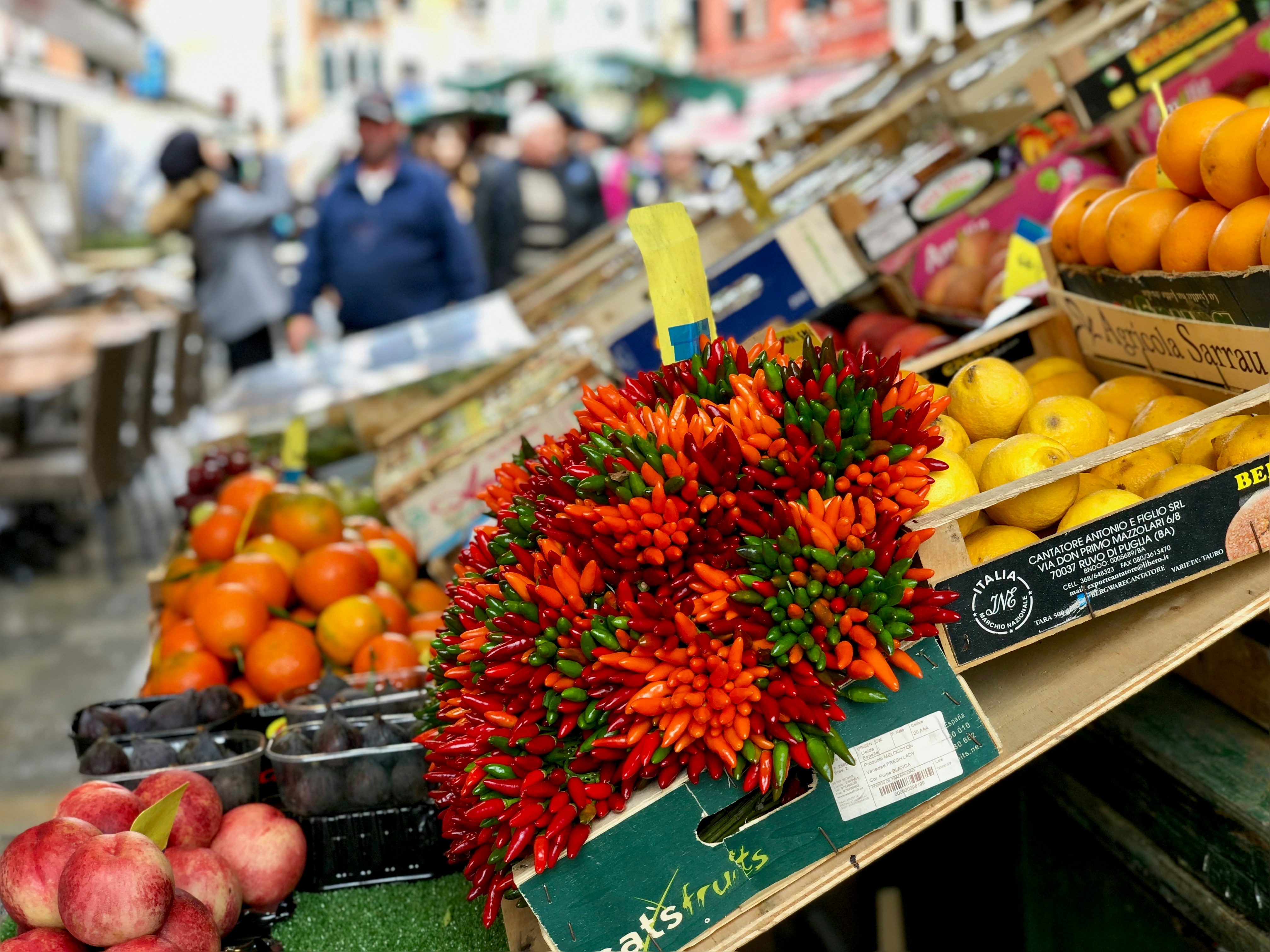 A bustling market scene featuring an array of colorful fruits, including bright red chilies, oranges, and apples, set against a lively background of shoppers. 