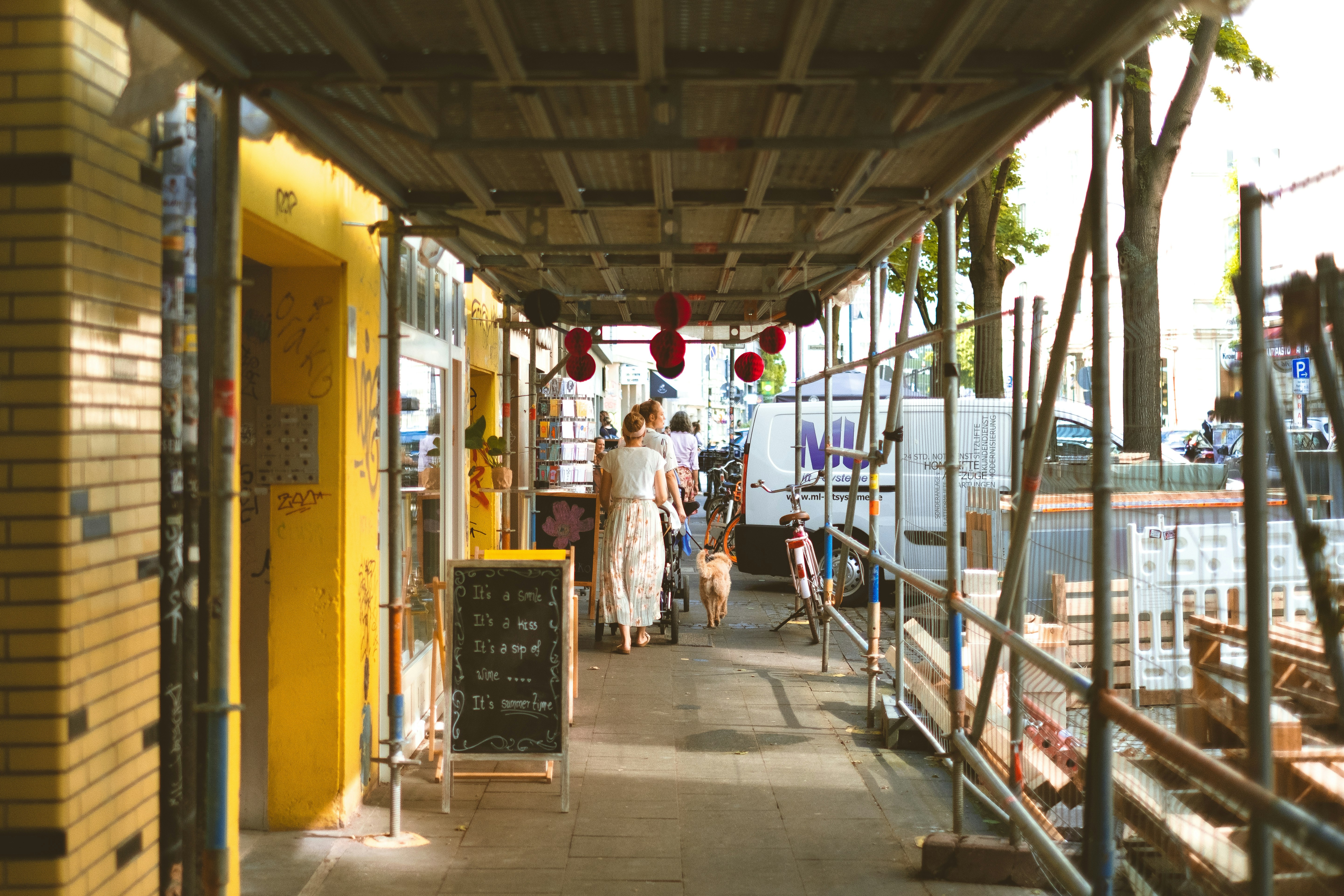 Two individuals walking under a construction canopy adorned with lanterns, flanked by vibrant storefronts. A dog accompanies them on the bustling sidewalk.