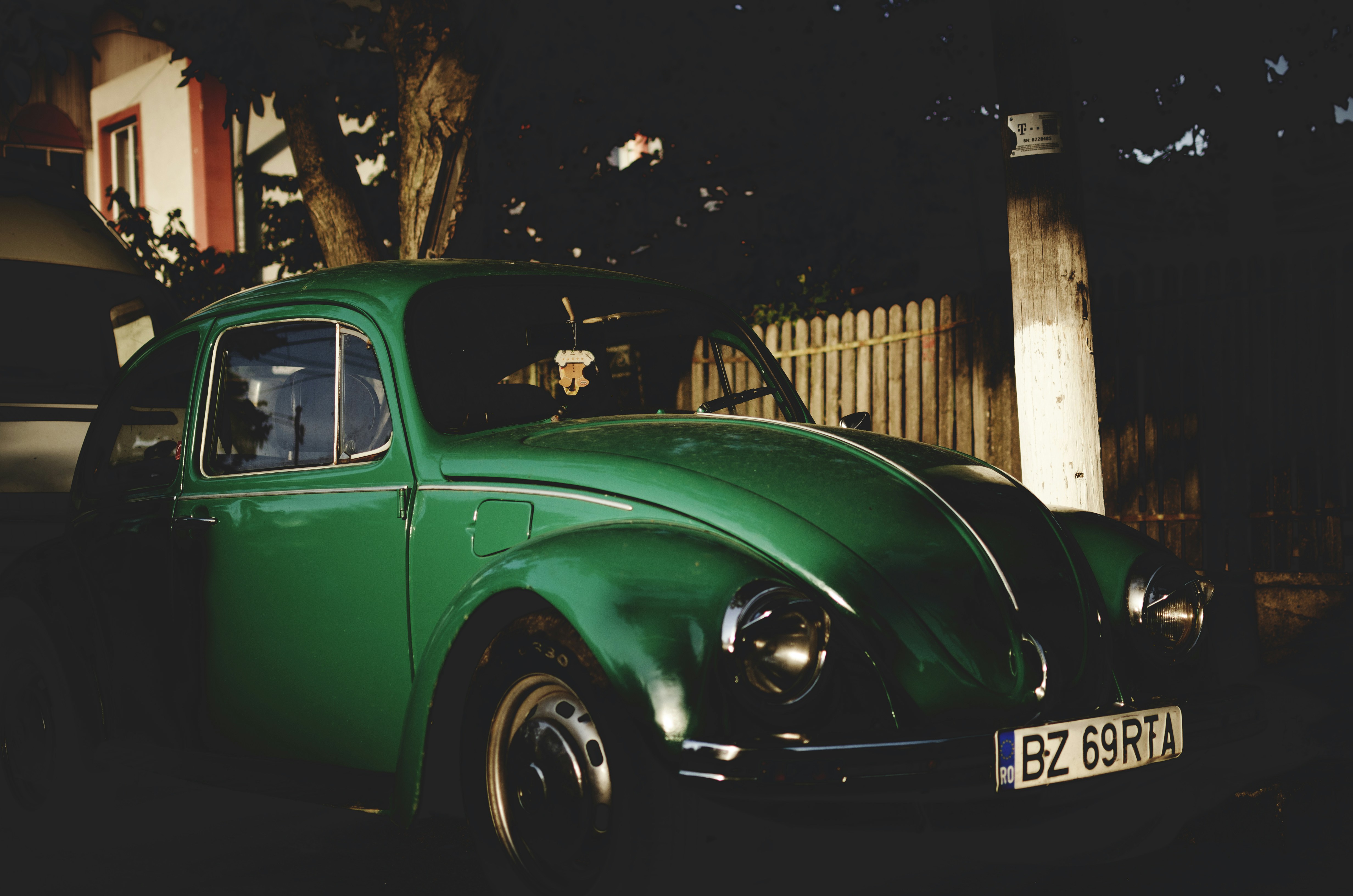 A classic green Volkswagen Beetle parked under soft street lighting, surrounded by trees and a wooden fence.