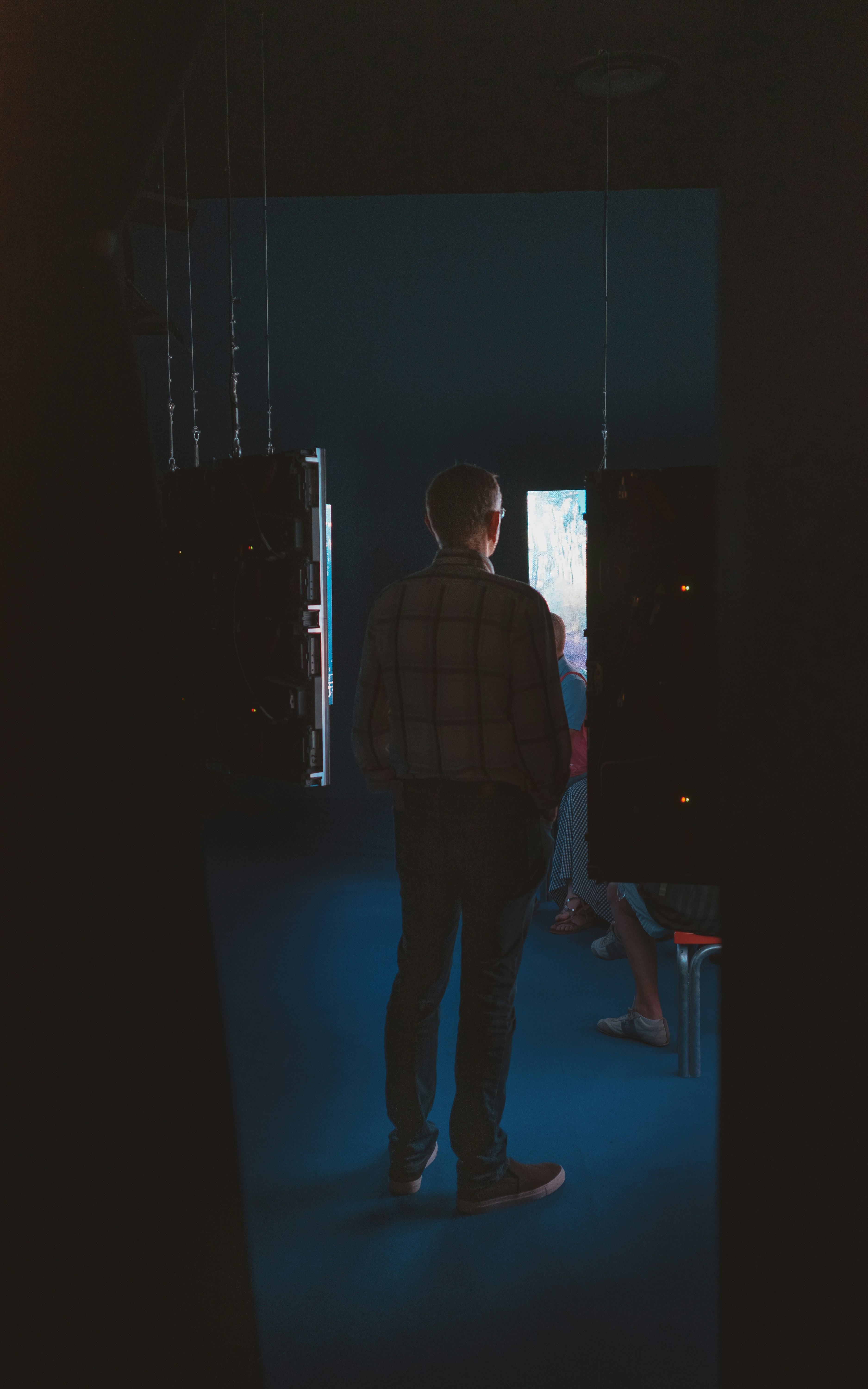 Blue Threshold: A Back-Turned Figure in a Dim Studio Framed by Hanging Monitors