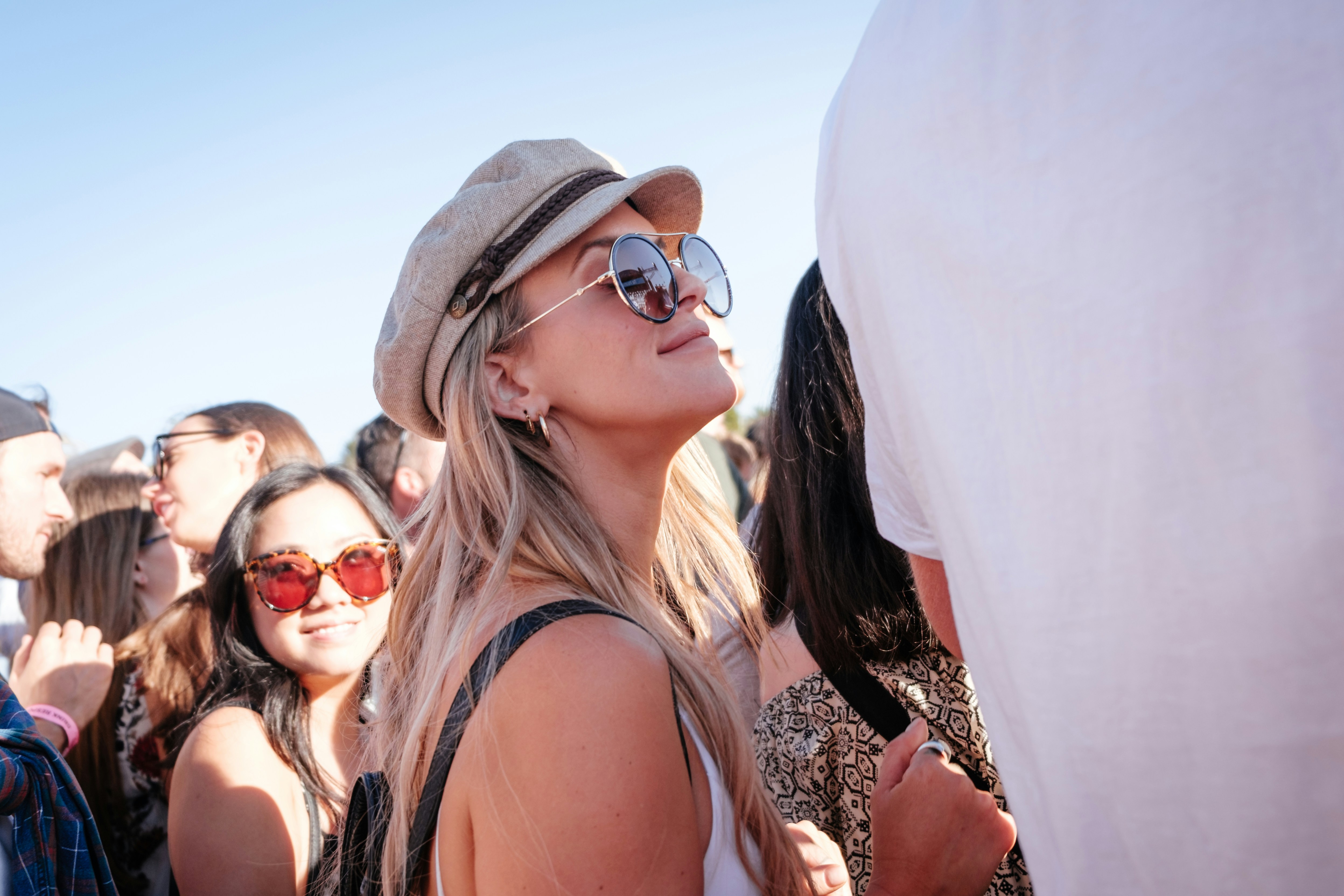 Festival-goer with stylish sunglasses and a cap, surrounded by a lively crowd under a clear blue sky.