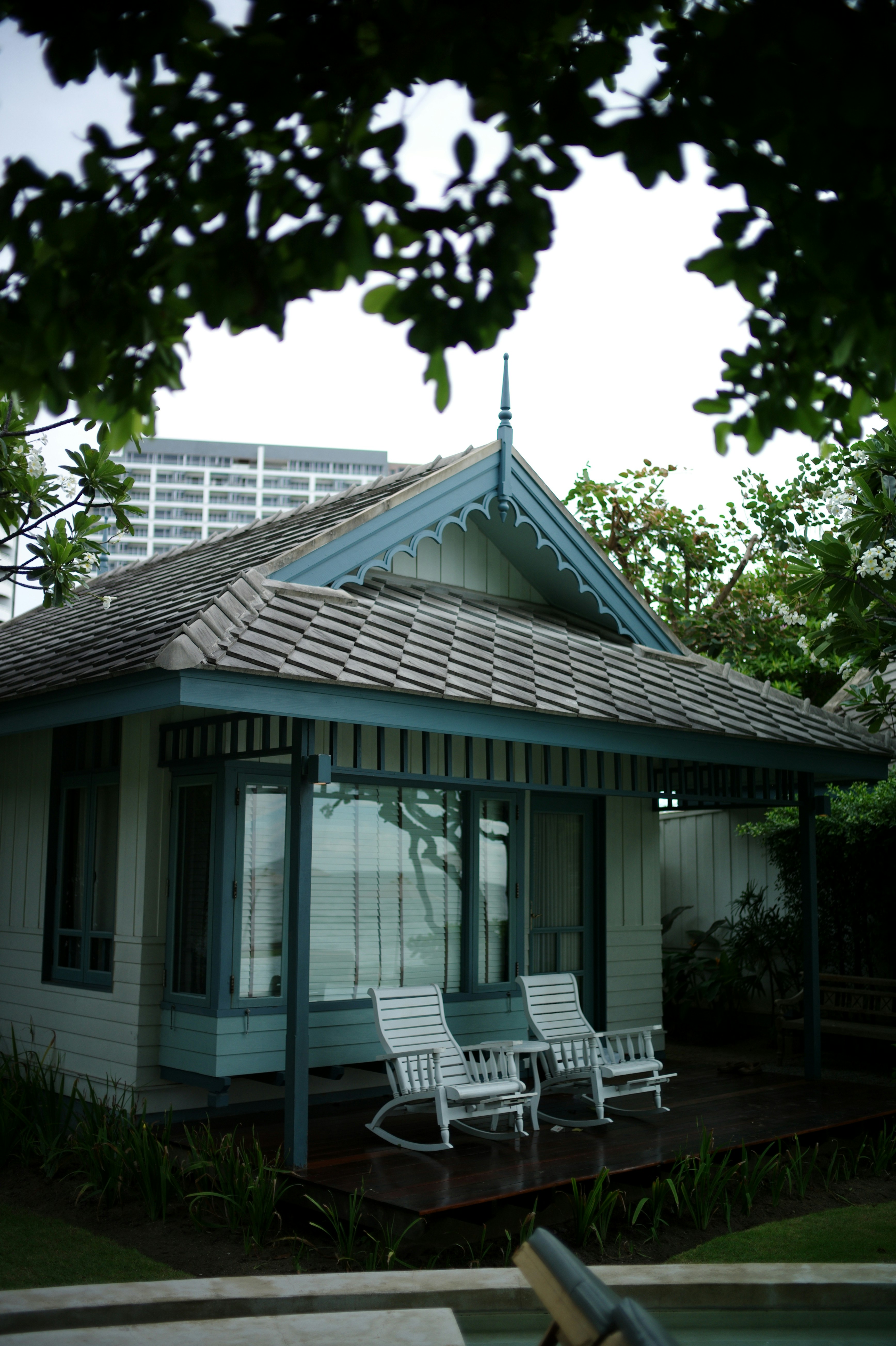 Charming coastal cottage with blue accents and rocking chairs, surrounded by lush greenery. A tranquil retreat against a backdrop of modern buildings.