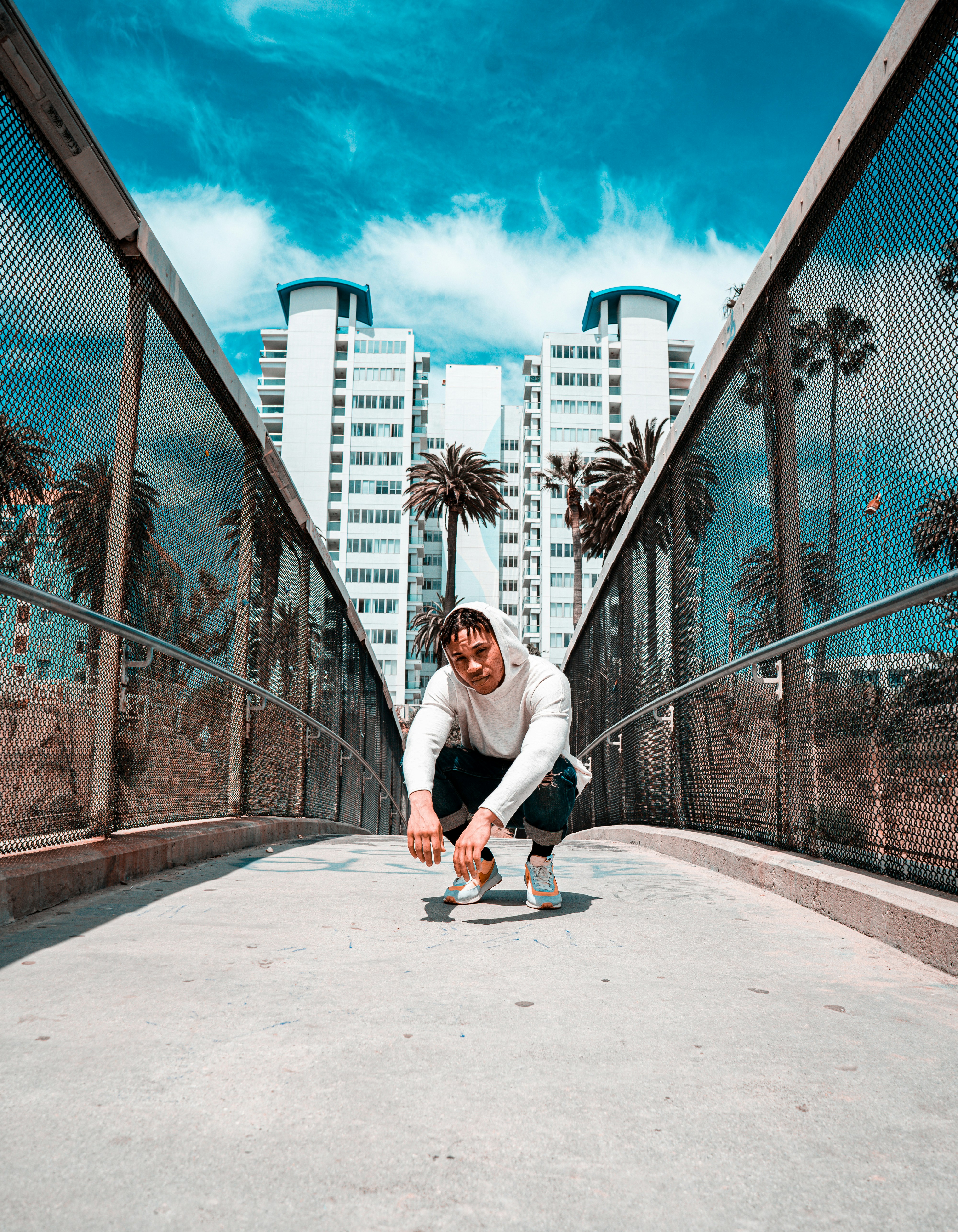 Young man crouching on a walkway, framed by palm trees and modern buildings under a vibrant blue sky.
