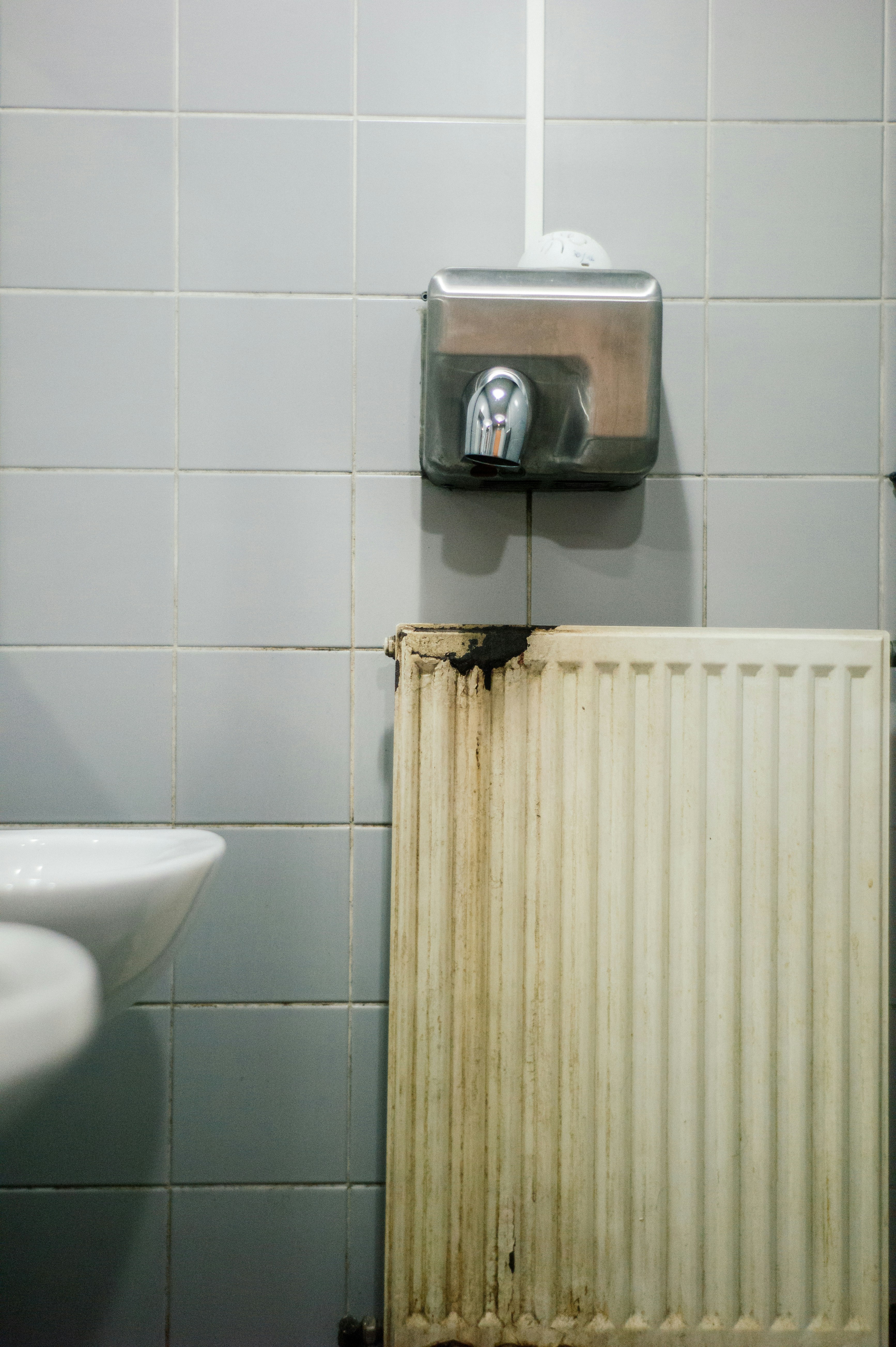 Metallic hand dryer mounted above a stained radiator in a utilitarian bathroom setting.
