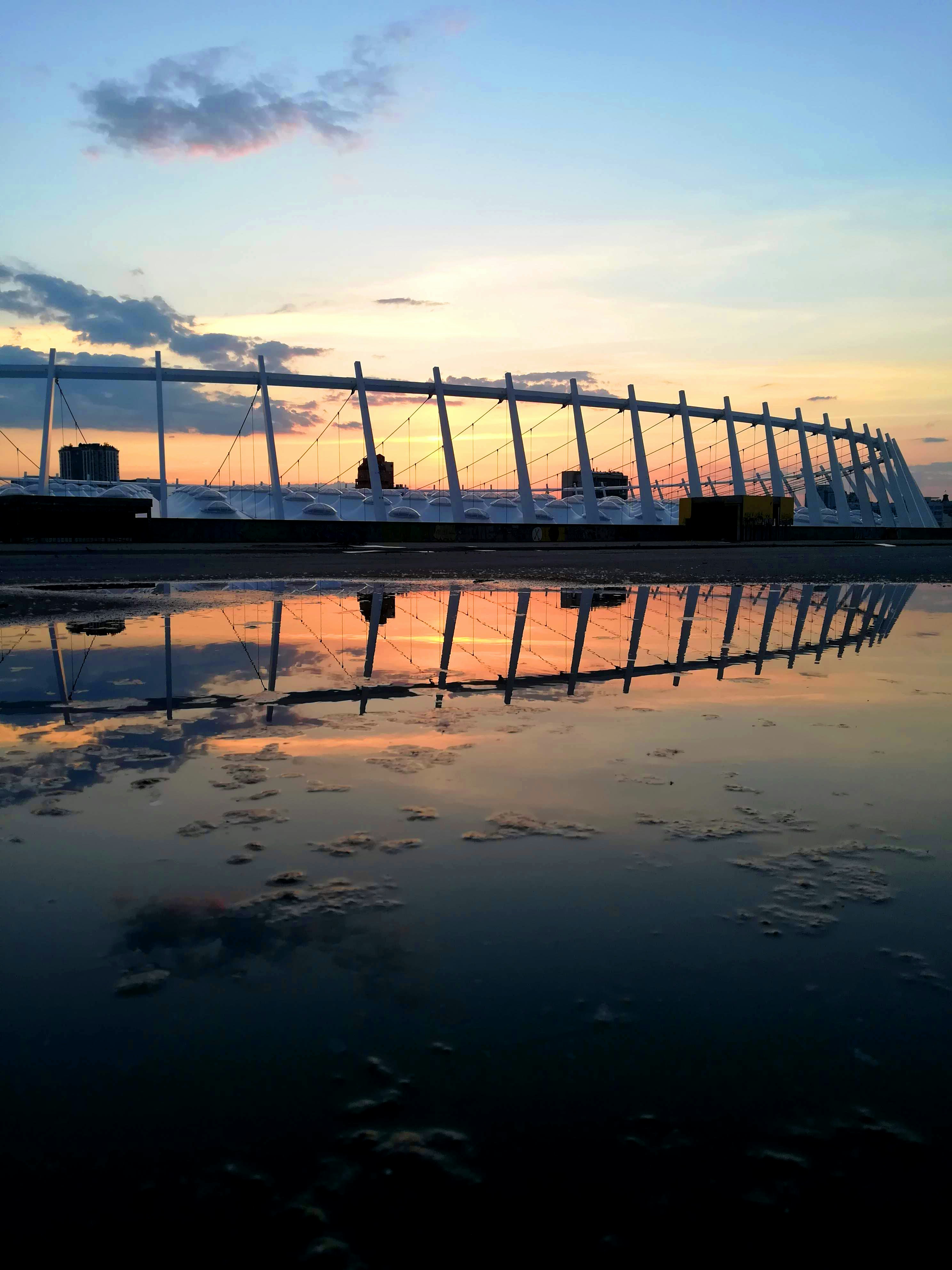 Modern architectural bridge reflecting in a puddle at sunset, with vibrant hues illuminating the sky.