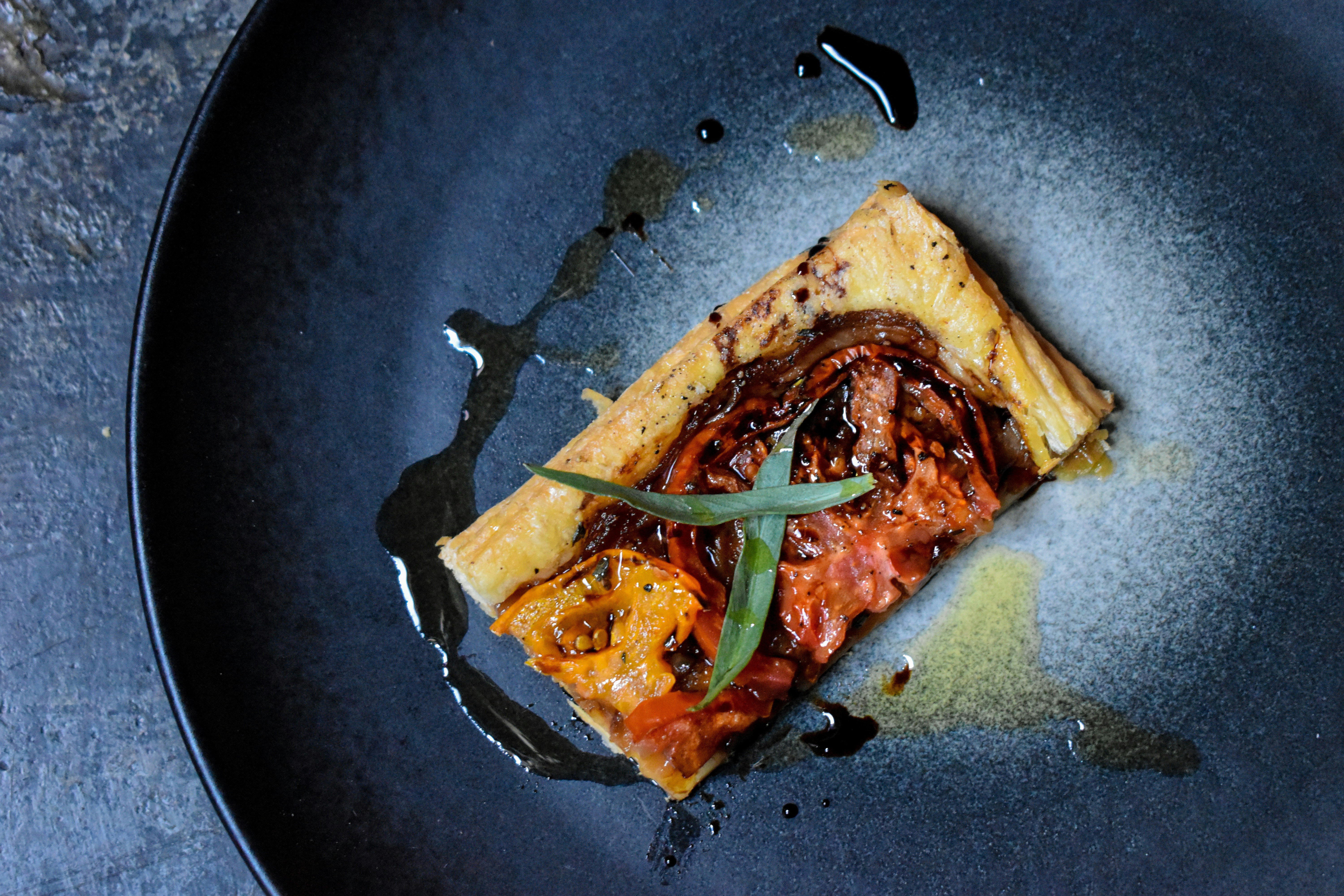 A beautifully plated tart featuring roasted vegetables and herbs, drizzled with olive oil on a dark plate.
