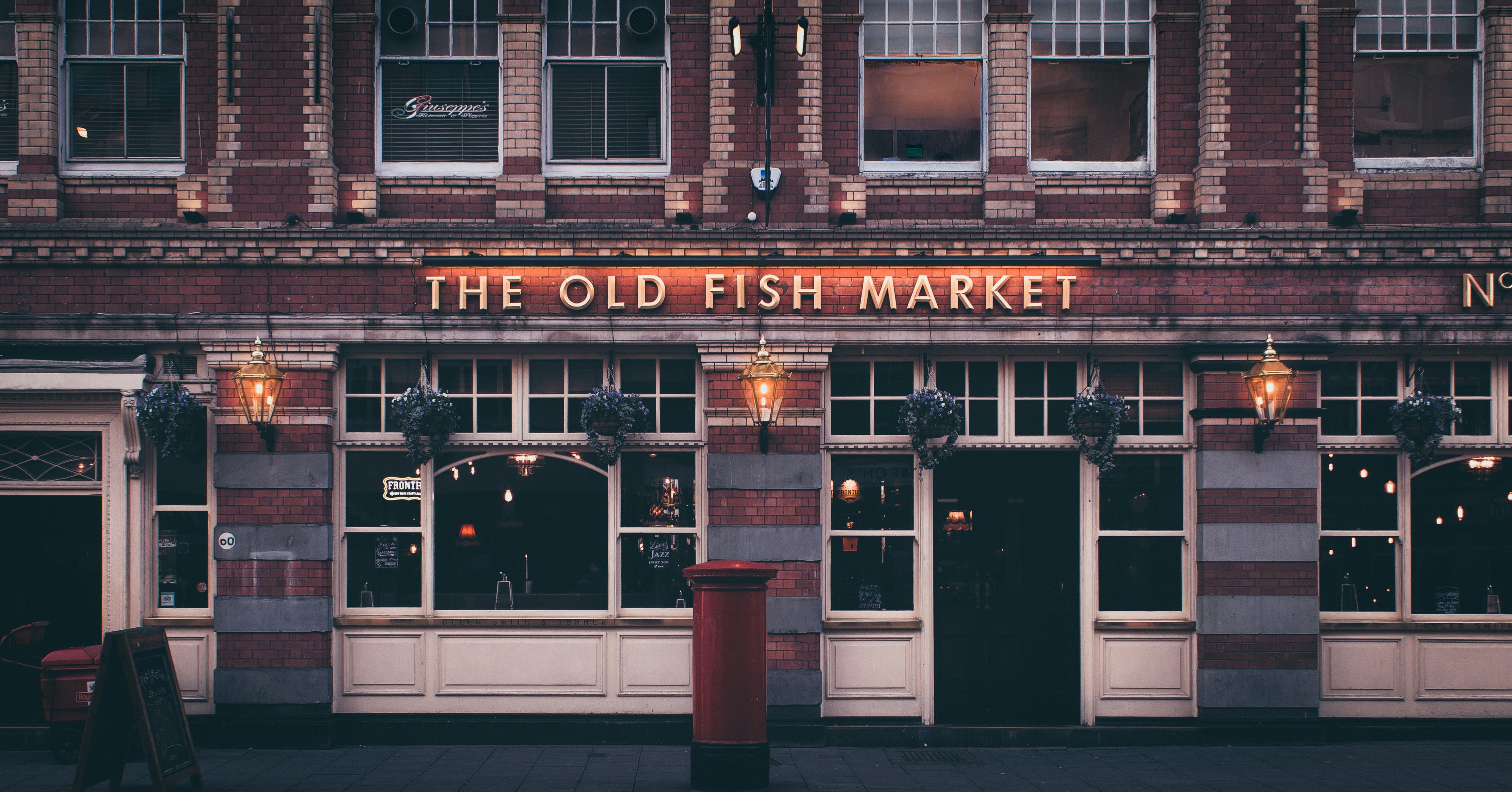 Historic storefront of The Old Fish Market showcasing vintage architecture and warm lighting. The inviting entrance features decorative lanterns and lush greenery.