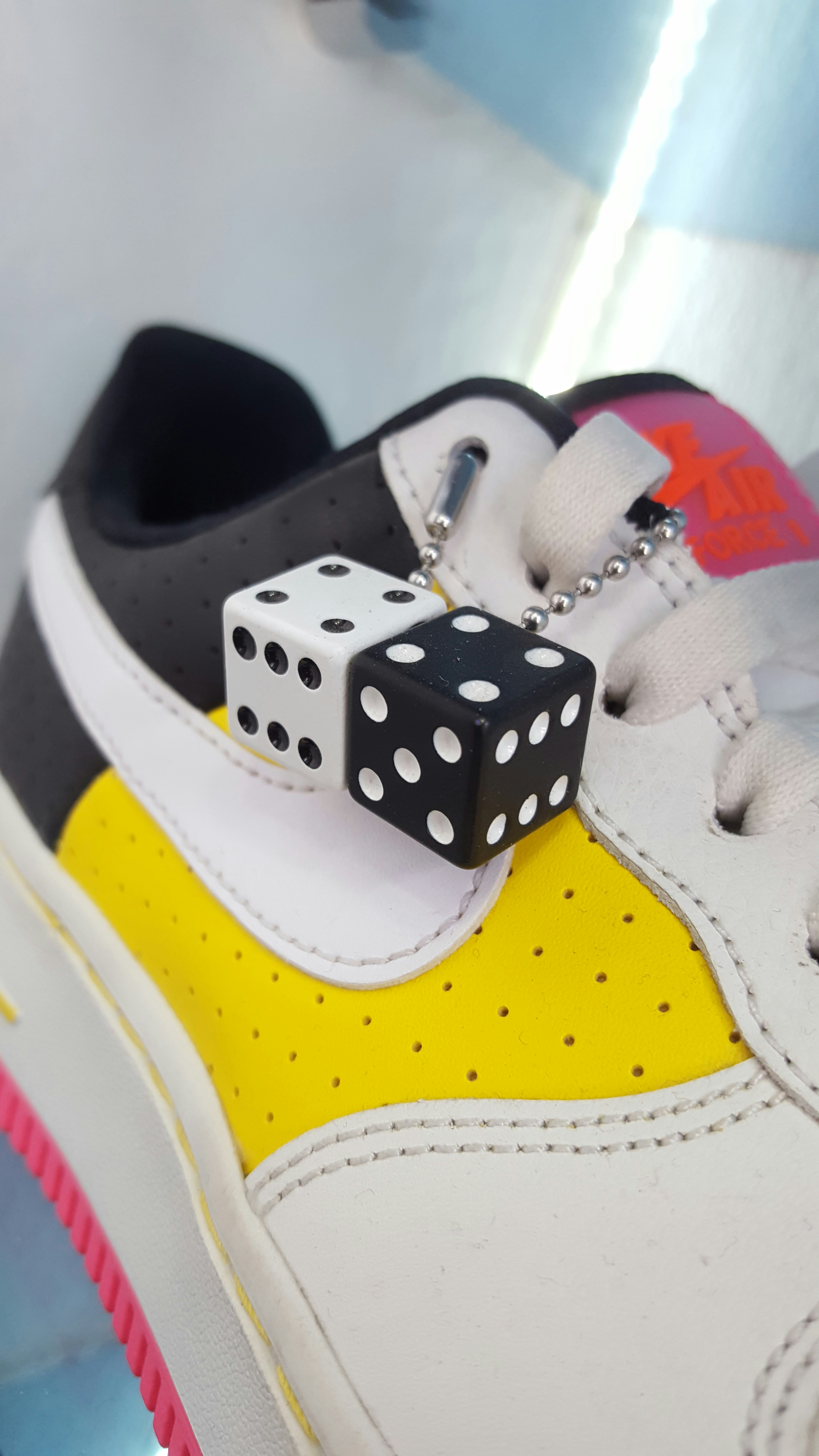 Close-up of matching white and black sneakers with yellow accents, featuring a pair of unique dice attached to the laces via a small chain. The scene emphasizes texture and color on the shoe surface.