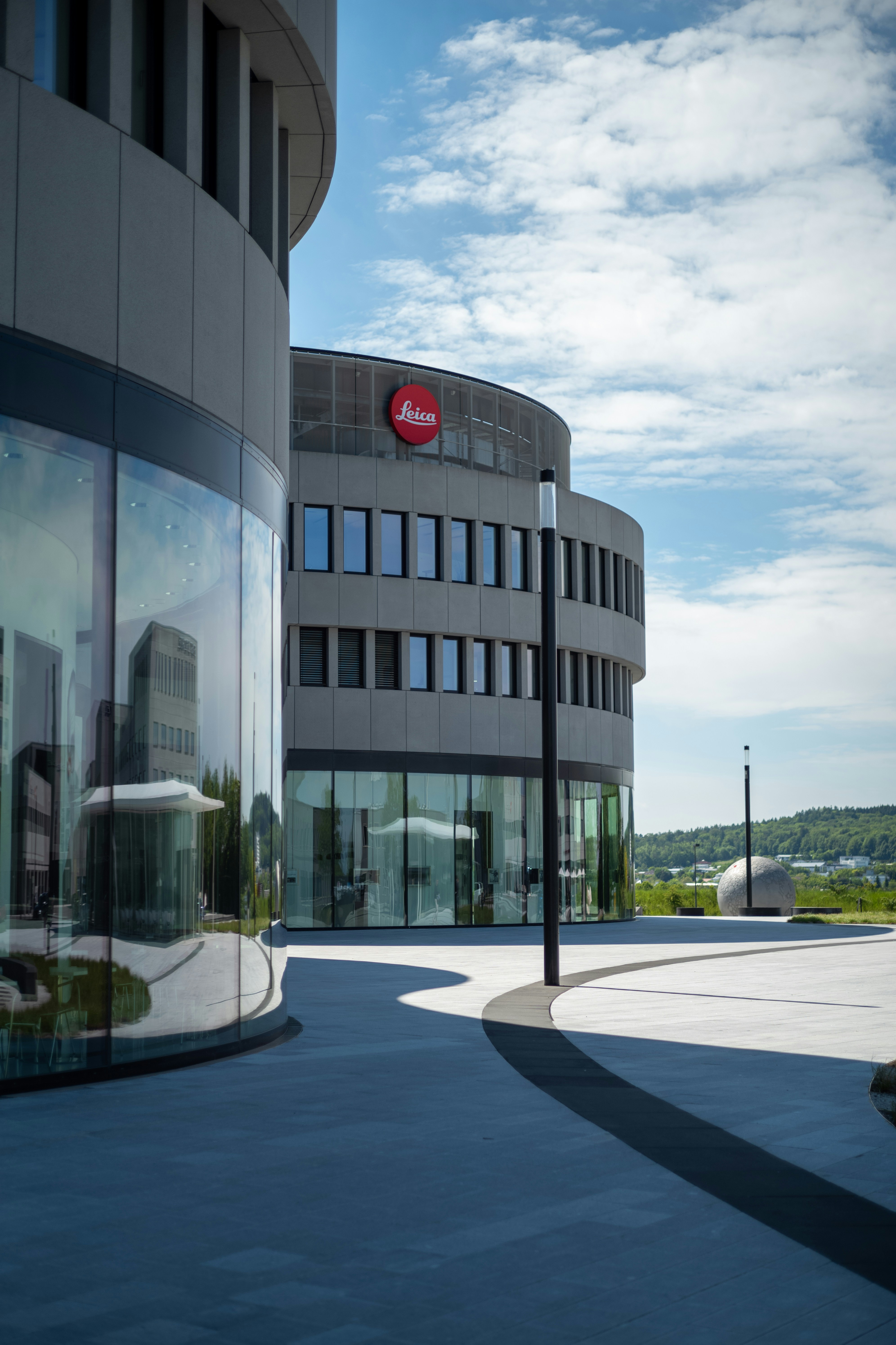 Curved glass facade reflecting the sky beside the cylindrical Leica headquarters under a partly cloudy sky.