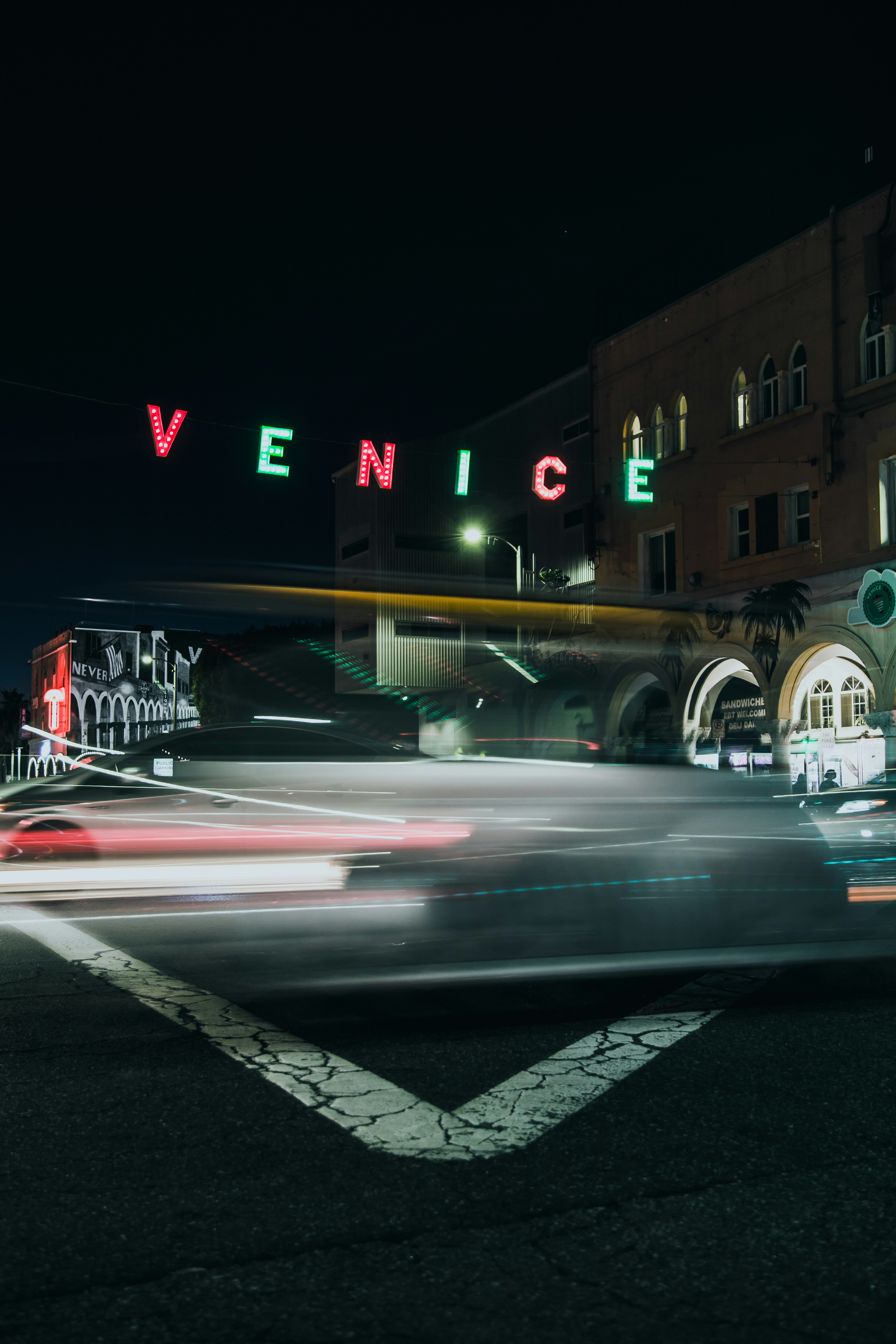 Venice Neon Over a Night Street: Long-Exposure Trails Beneath a Glowing Sign