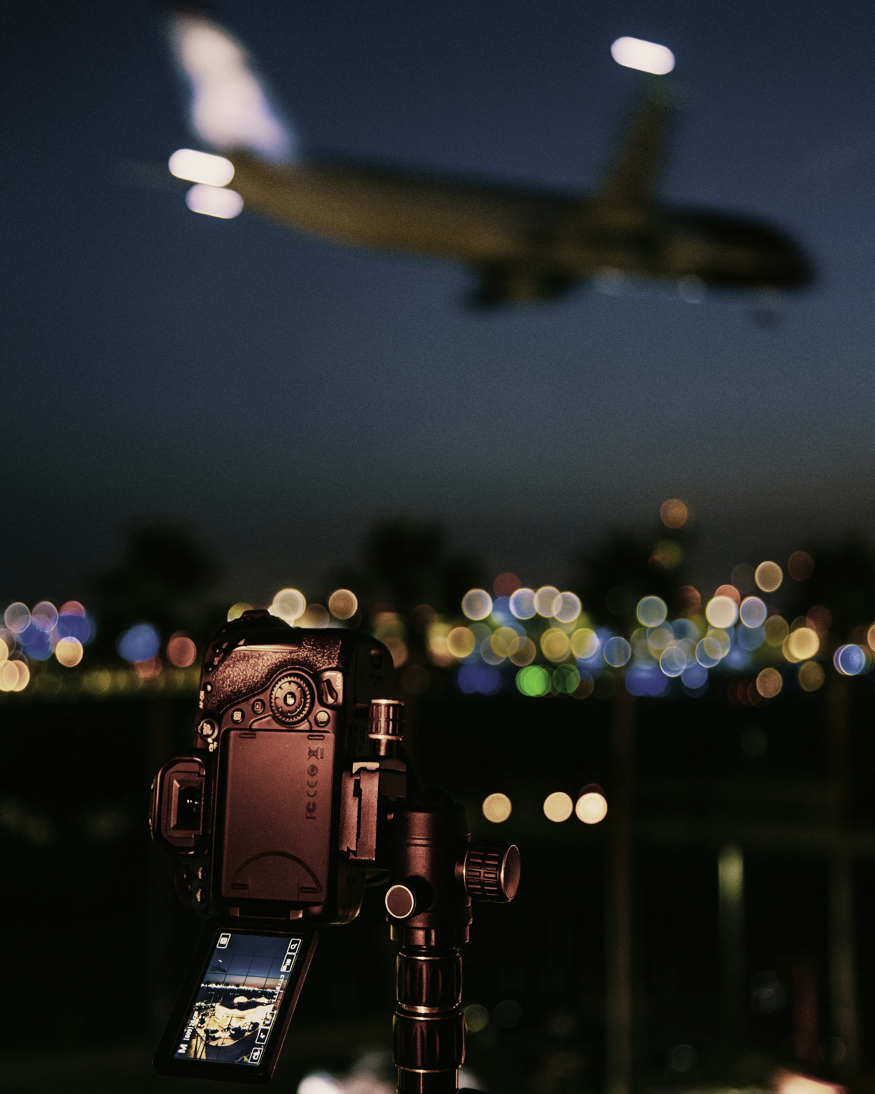 Camera positioned on a tripod capturing an aircraft in motion against a backdrop of vibrant bokeh lights at dusk.