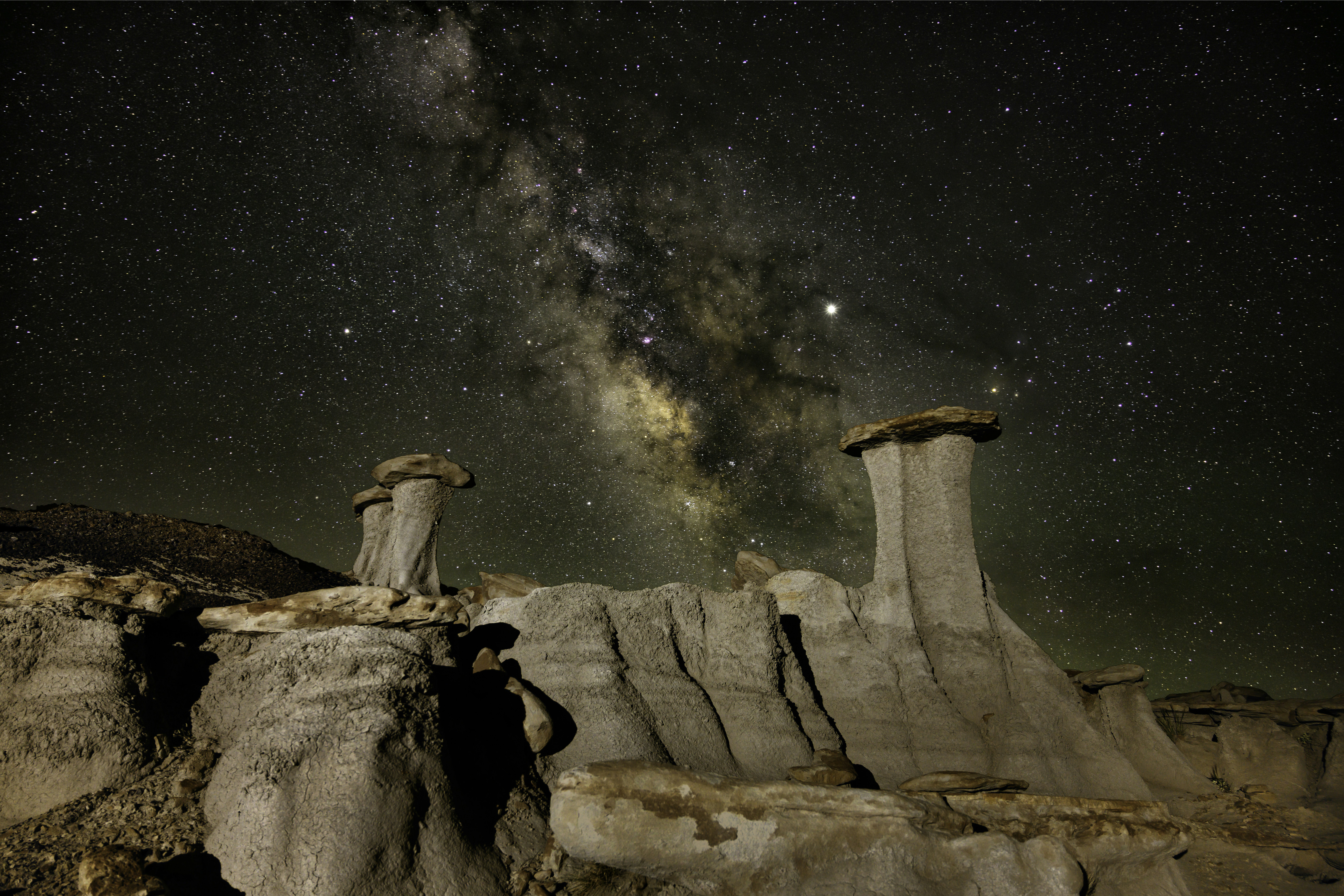 Night photograph of the Milky Way arcing over eroded desert hoodoos and rugged foreground. The celestial arc stands against the sculpted rock landscape, guiding the viewer through the scene.