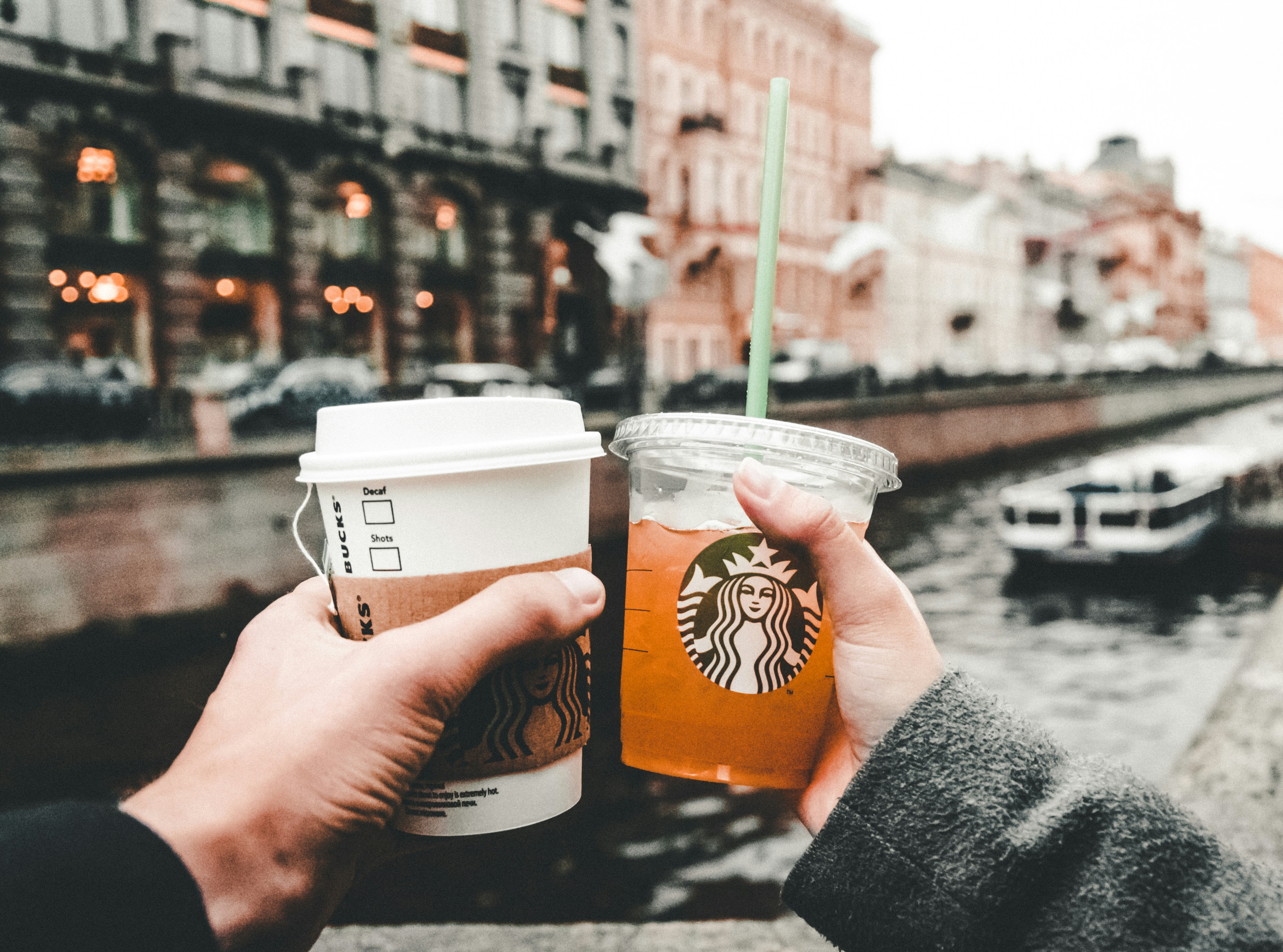 Two hands holding Starbucks drinks against a picturesque canal backdrop, capturing a moment of shared enjoyment.