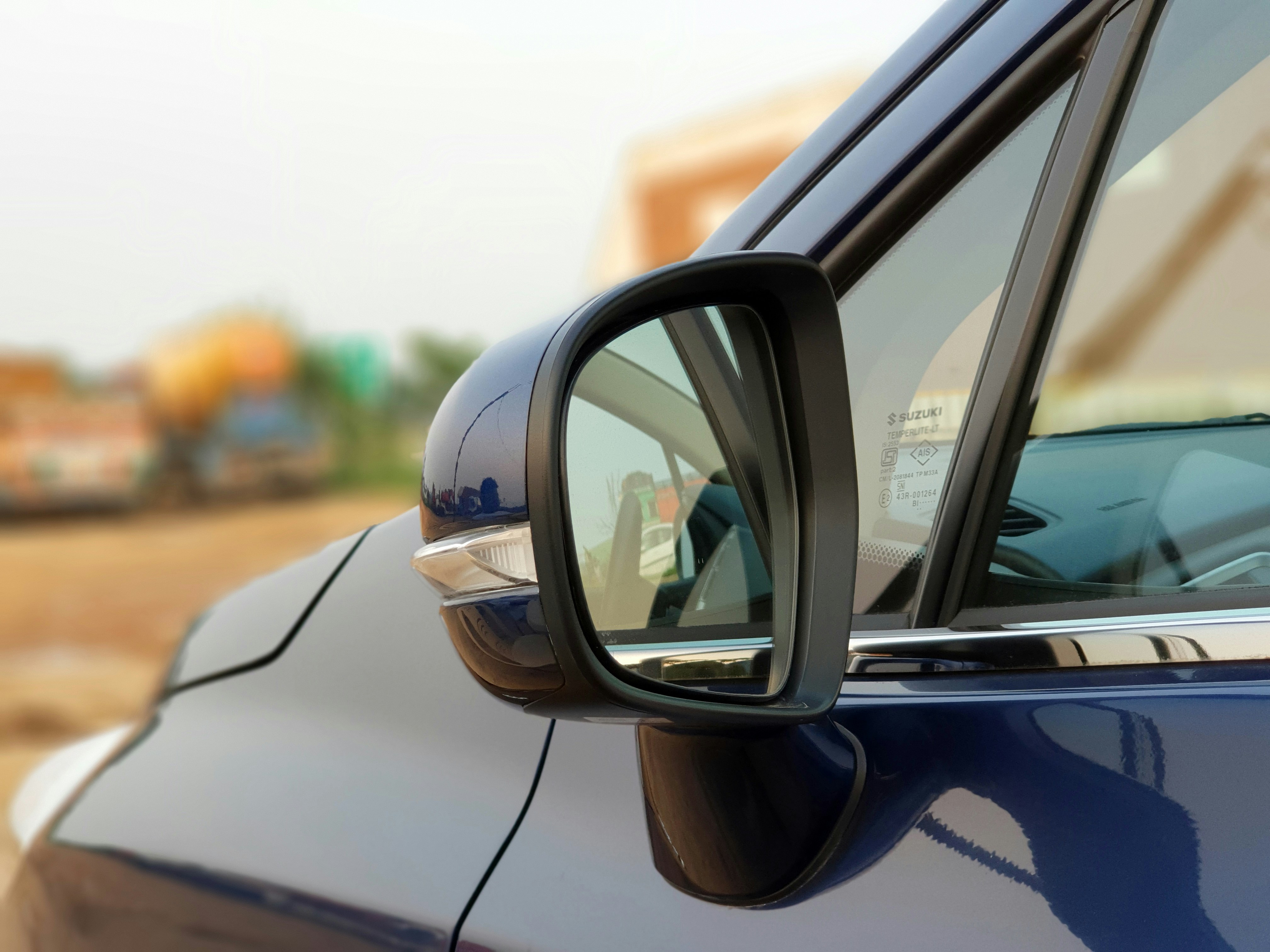Close-up of a car's side mirror reflecting the surroundings, showcasing the sleek design and details of the vehicle. The background features blurred shapes and colors, enhancing the focus on the mirror's reflection.