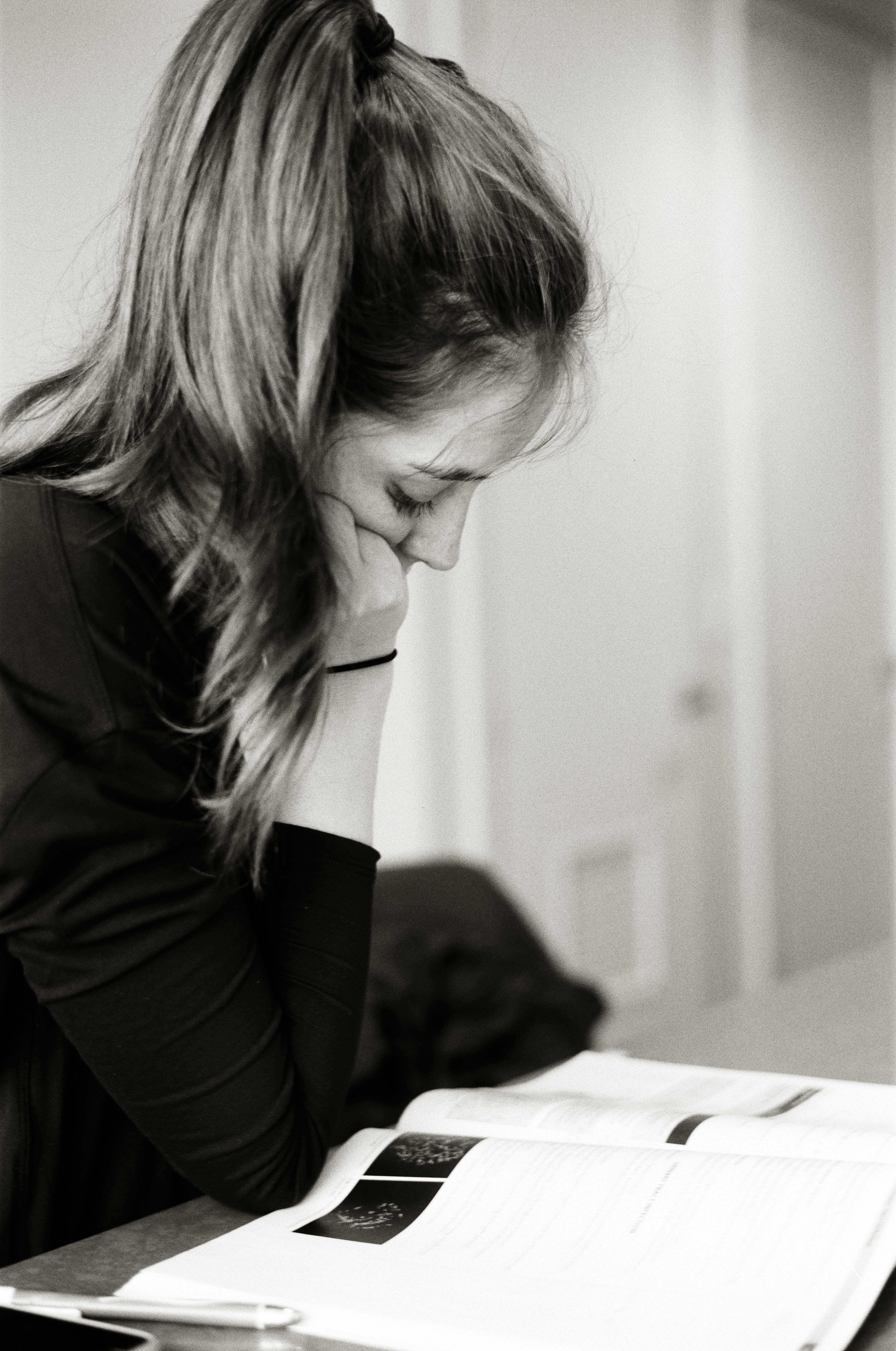 A young woman with long hair resting her chin on her hand, deeply focused on an open book on a table.
