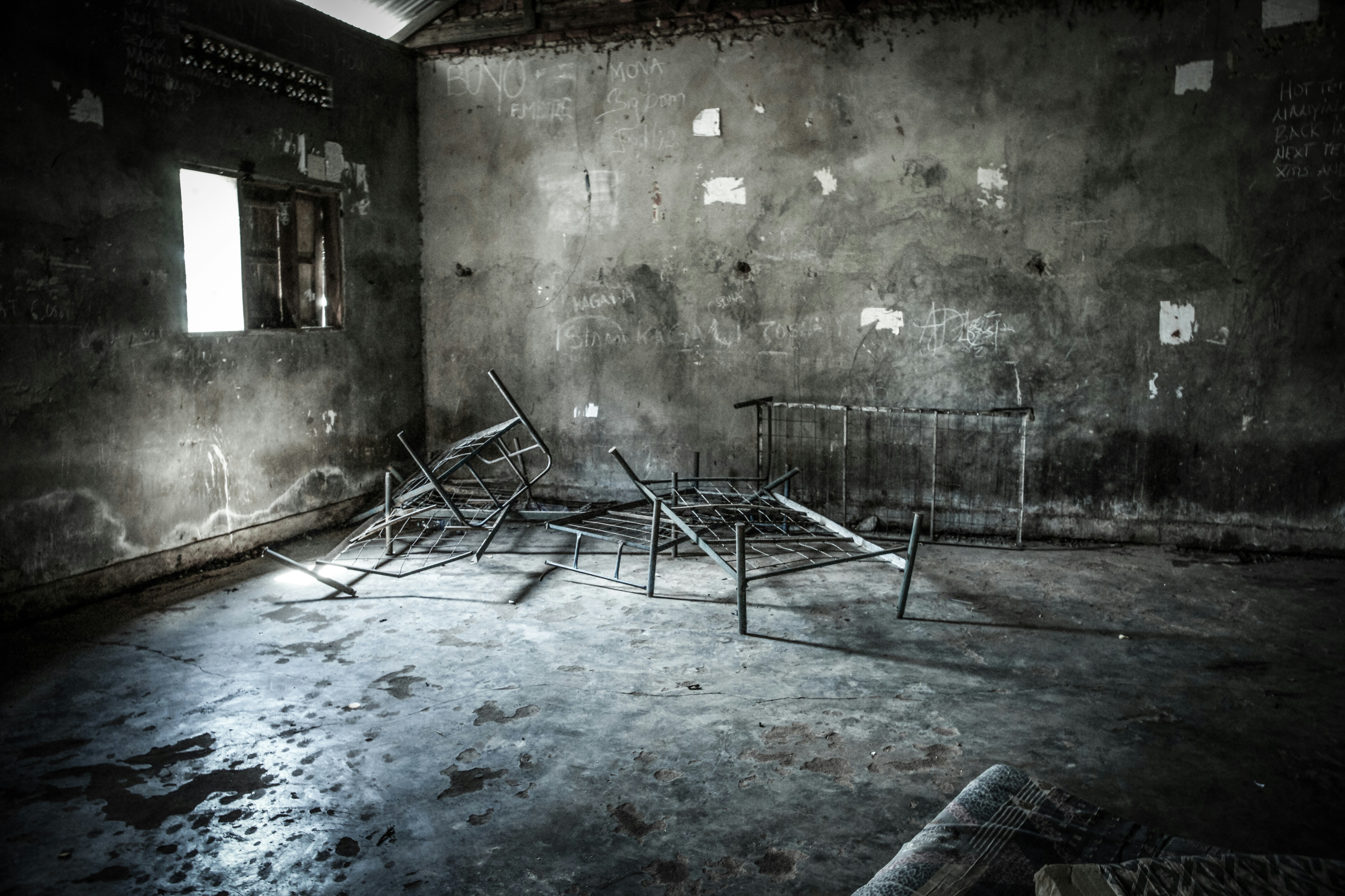 An abandoned room with scattered metal furniture and peeling walls, illuminated by soft light from a window. The scene evokes a sense of desolation.