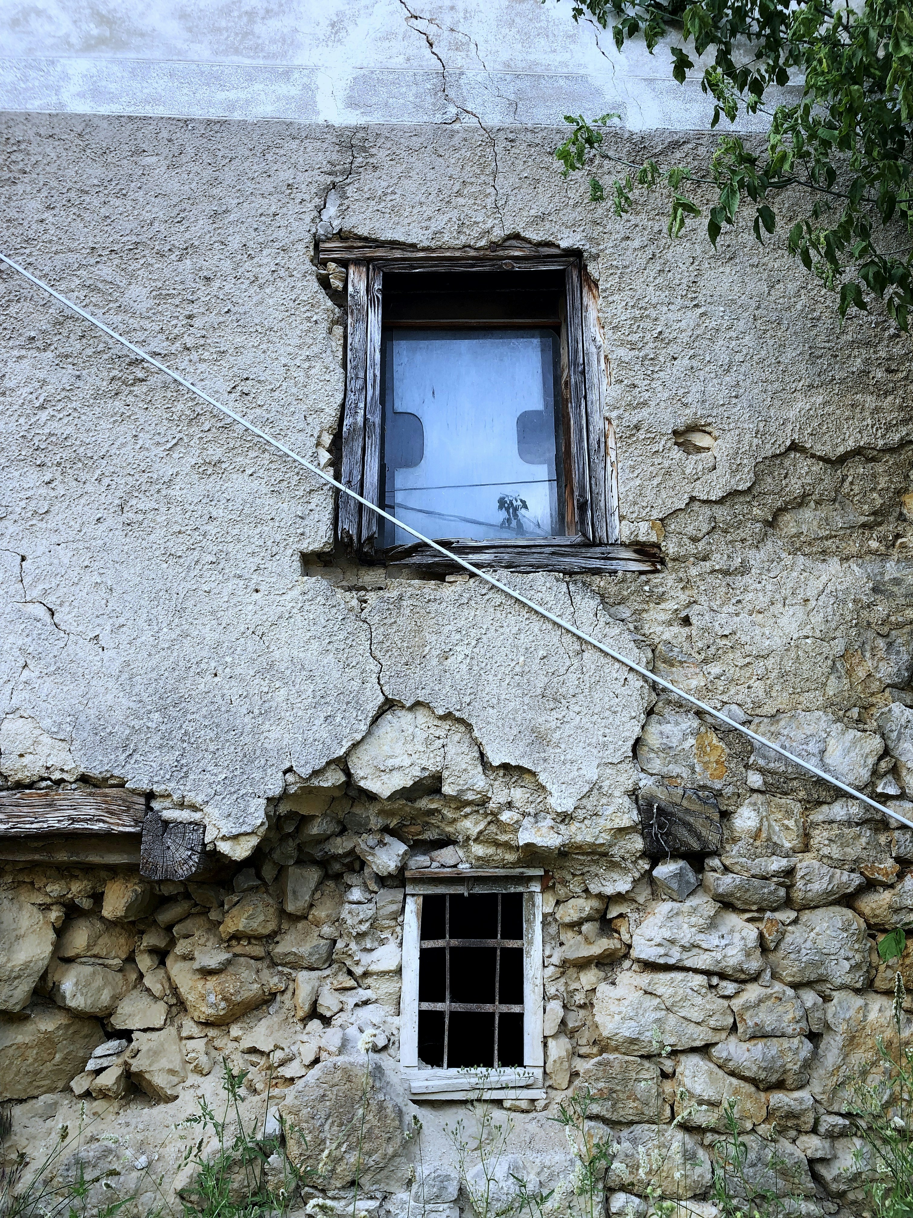 A weathered stone wall featuring two windows, one framed in rustic wood, revealing the passage of time. The texture of the wall contrasts with the greenery above.