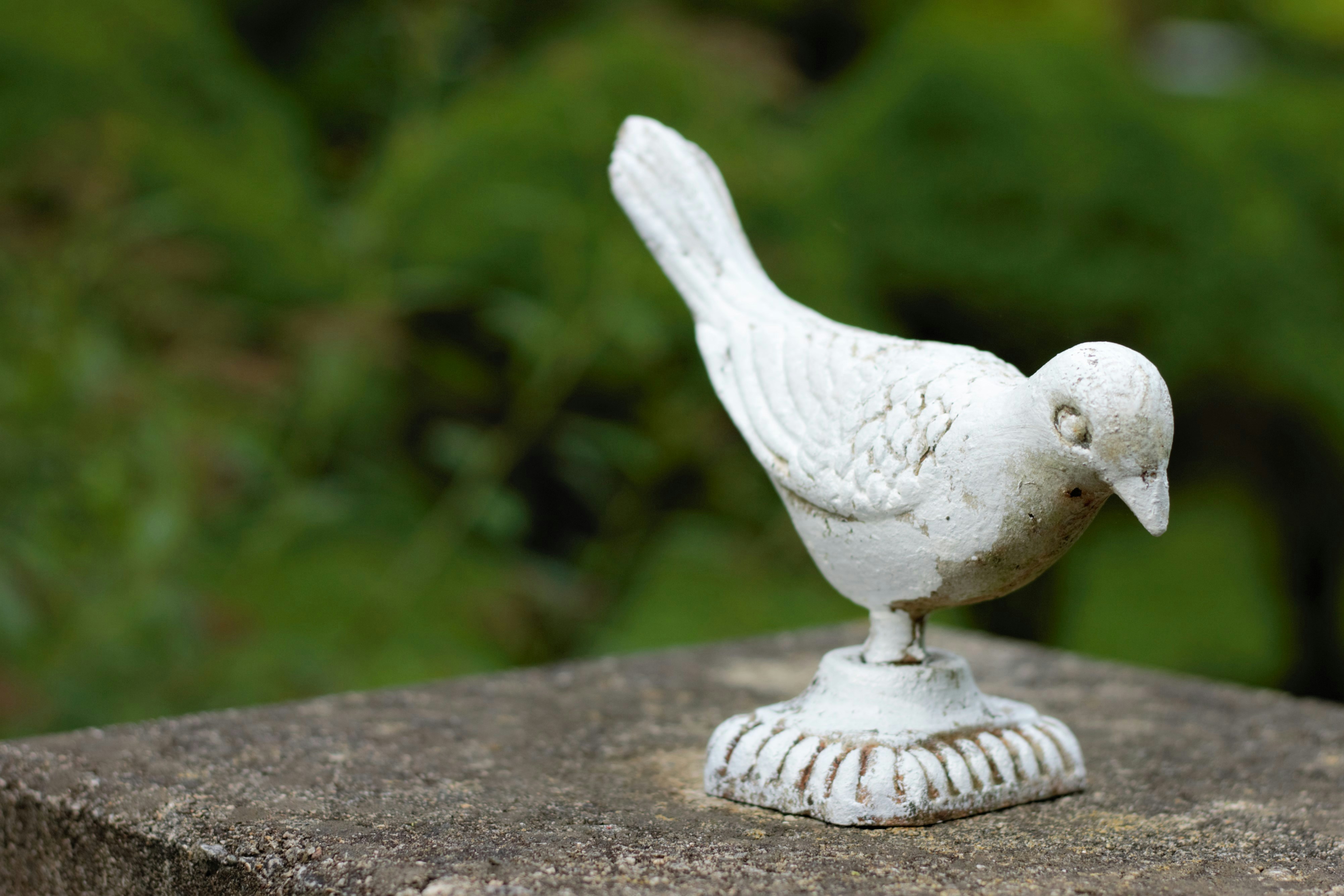 A cast iron bird sculpture perched on a stone base, surrounded by lush greenery. The intricate details of the bird contrast with the natural background.