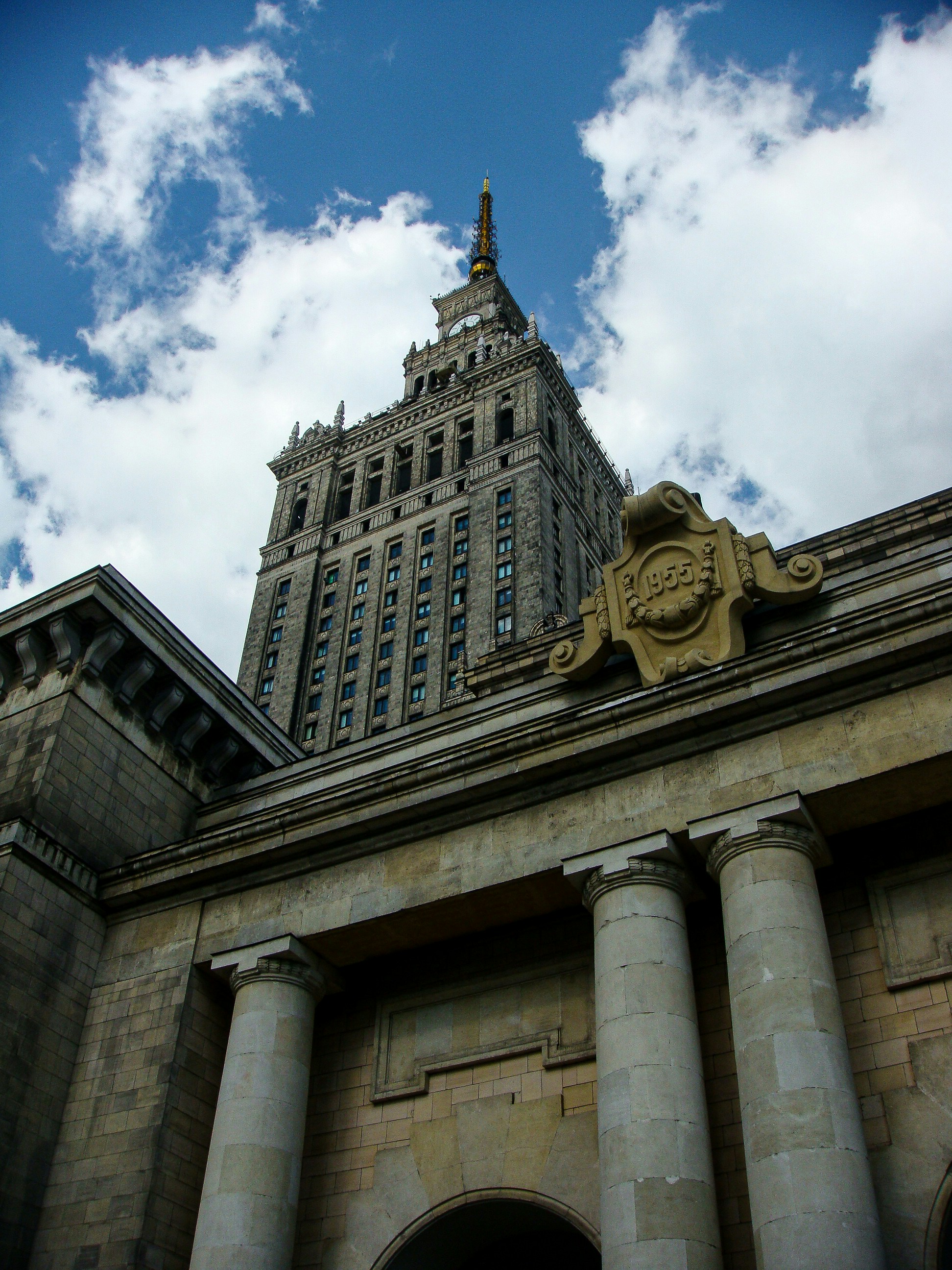 Angled view of a grand neoclassical facade with a towering spire, ornate crest, and stone columns against a bright blue sky.