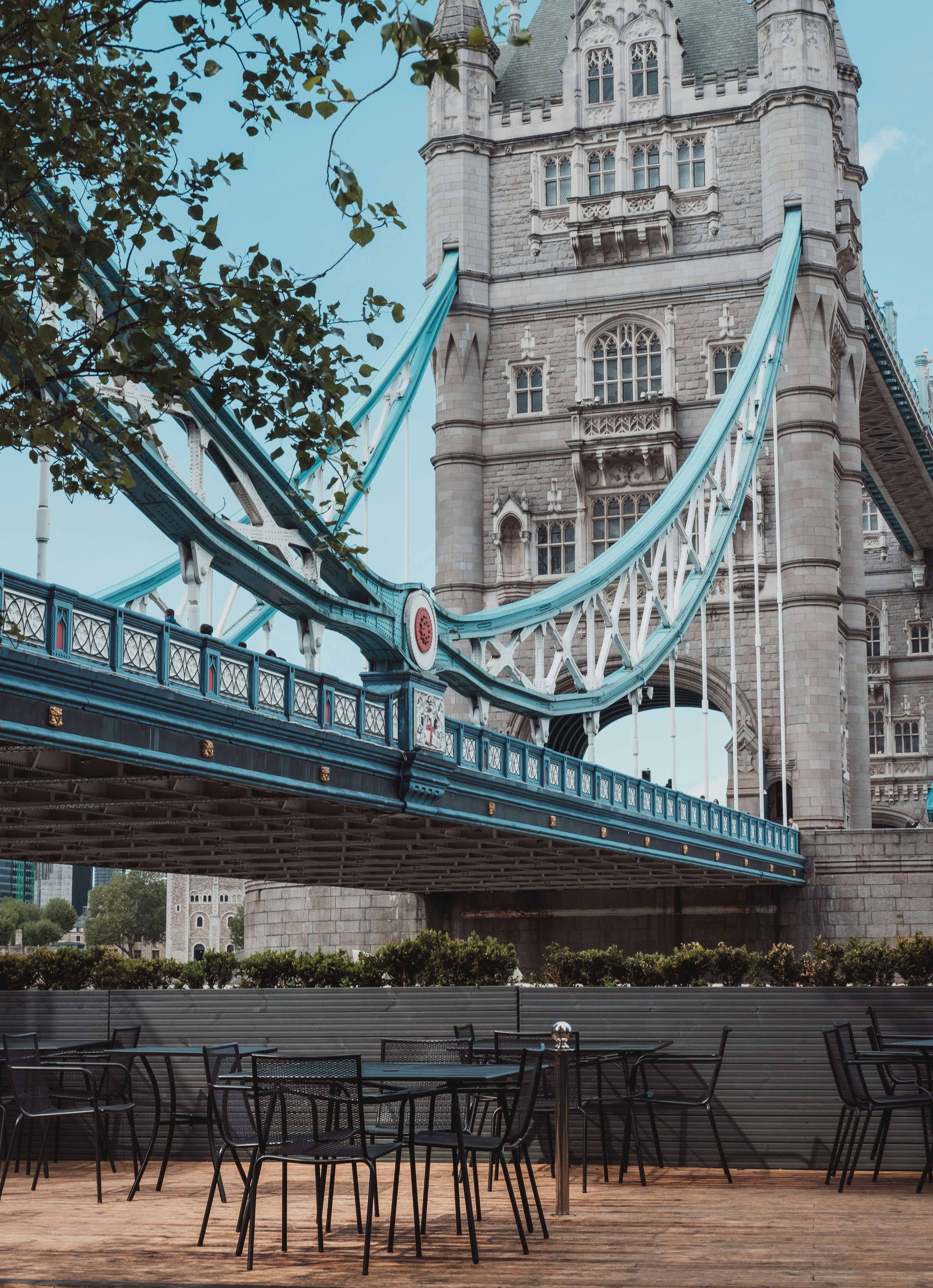 Tower Bridge stands majestically over the Thames, framed by lush greenery and an inviting riverside café. The architectural details highlight its iconic structure.
