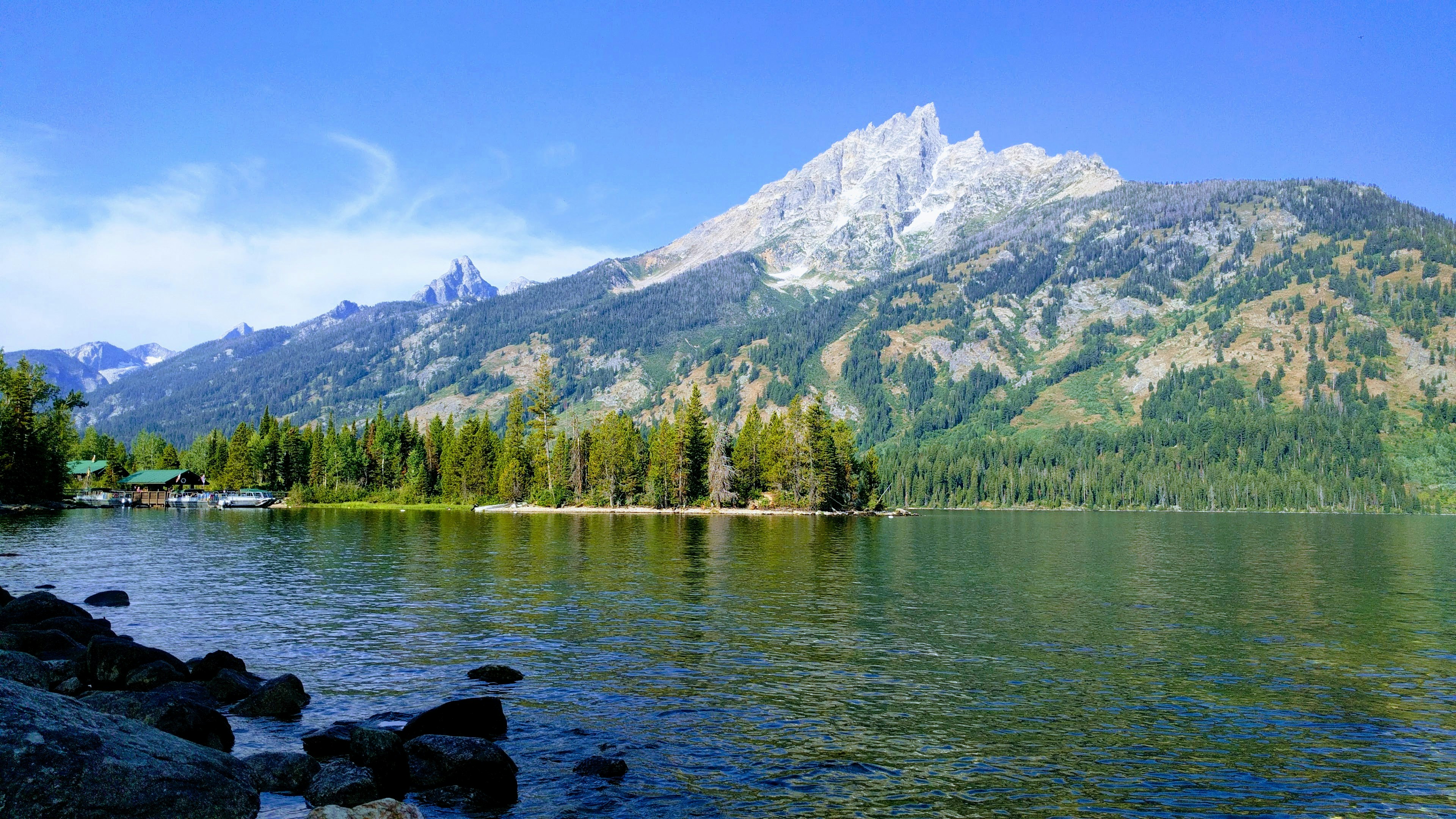 Mountain landscape with a clear lake reflecting the snow-capped peaks and surrounding trees under a bright blue sky.
