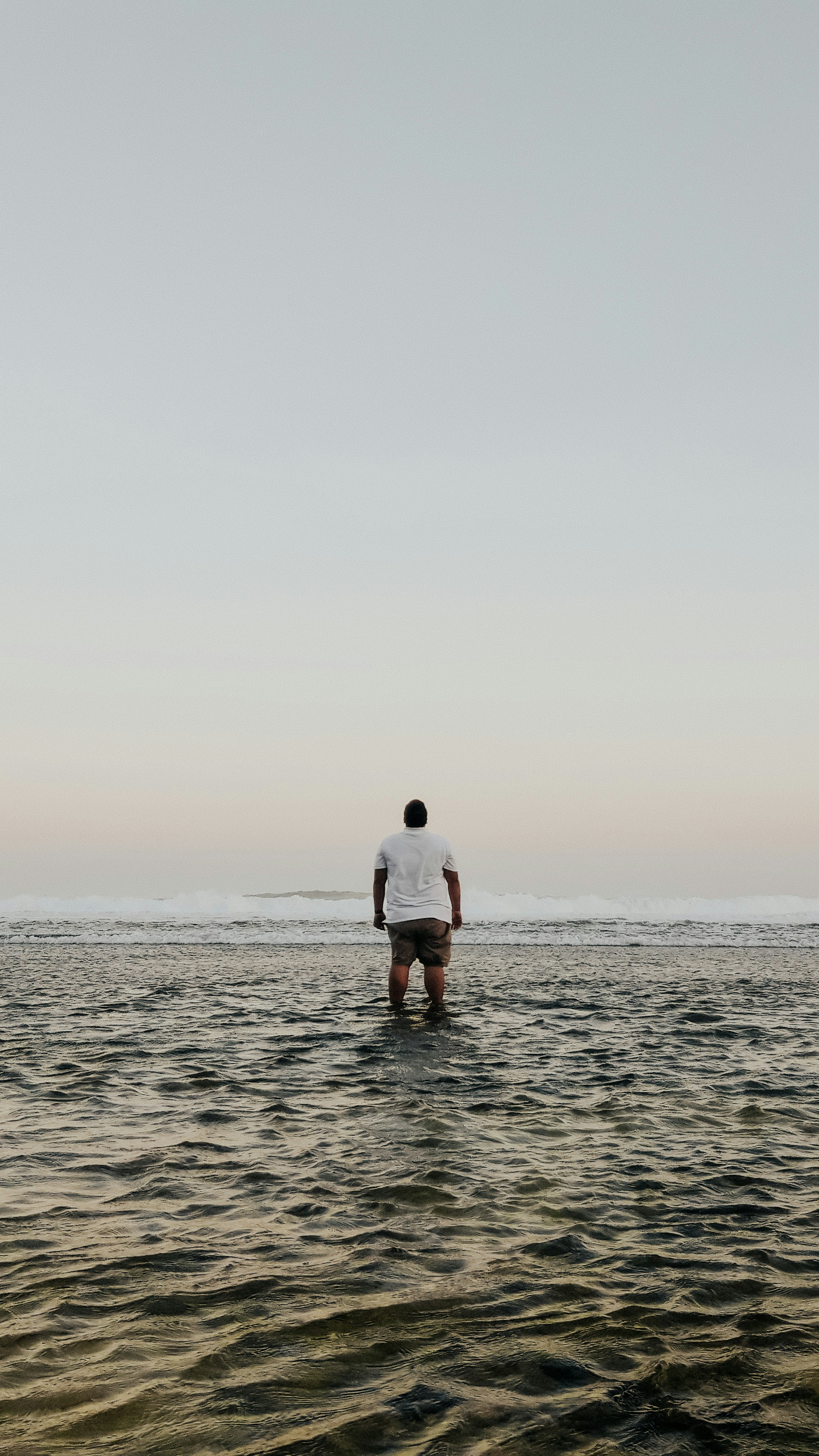 A lone figure stands in shallow water, gazing at the horizon where the sea meets the sky. The tranquil setting evokes a sense of introspection.