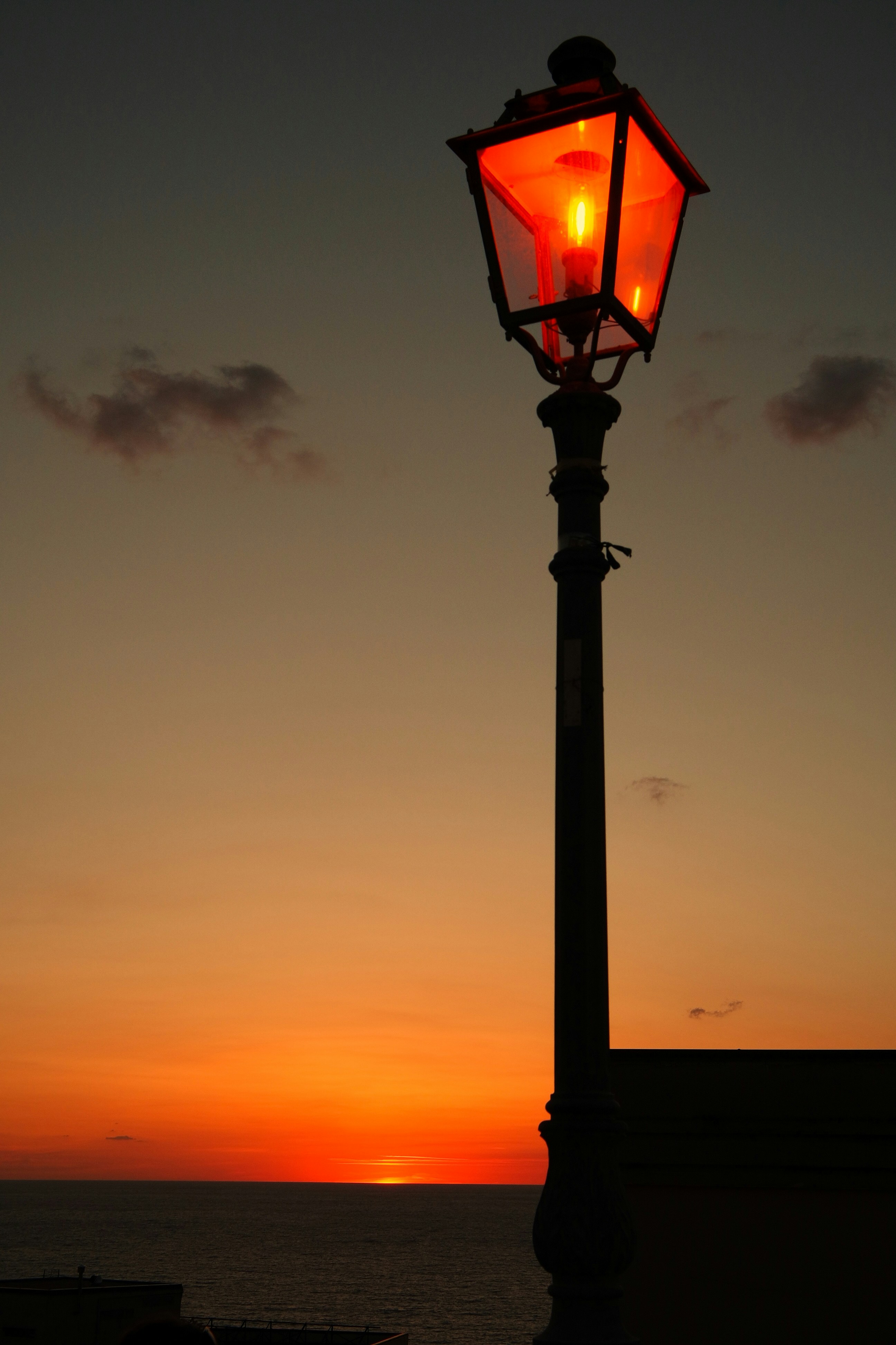 A vintage street lamp glowing warmly against a vibrant sunset over the ocean. The silhouette of the lamp contrasts beautifully with the colorful sky.