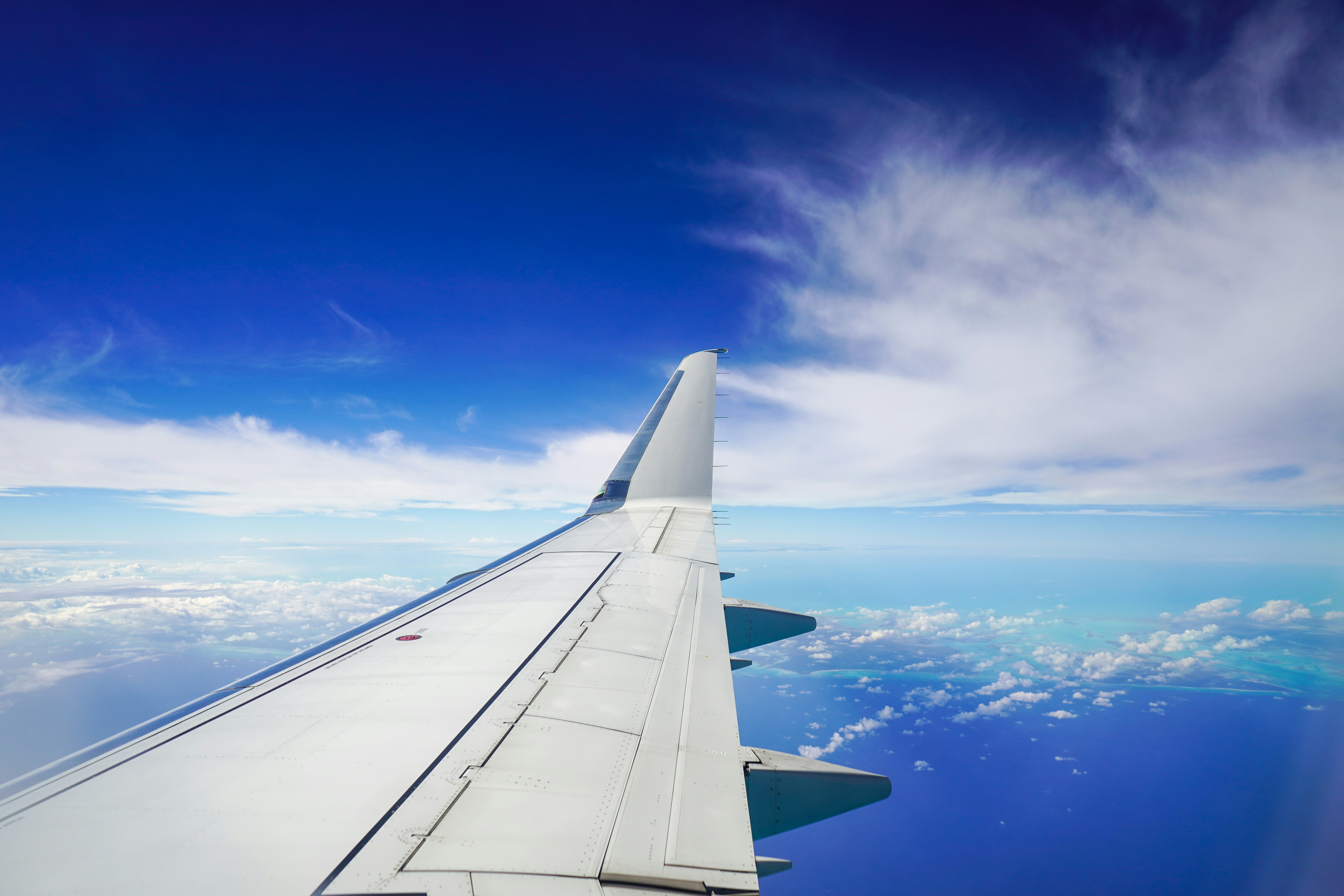 Airplane wing extending into a vast blue sky with wispy clouds and ocean below.