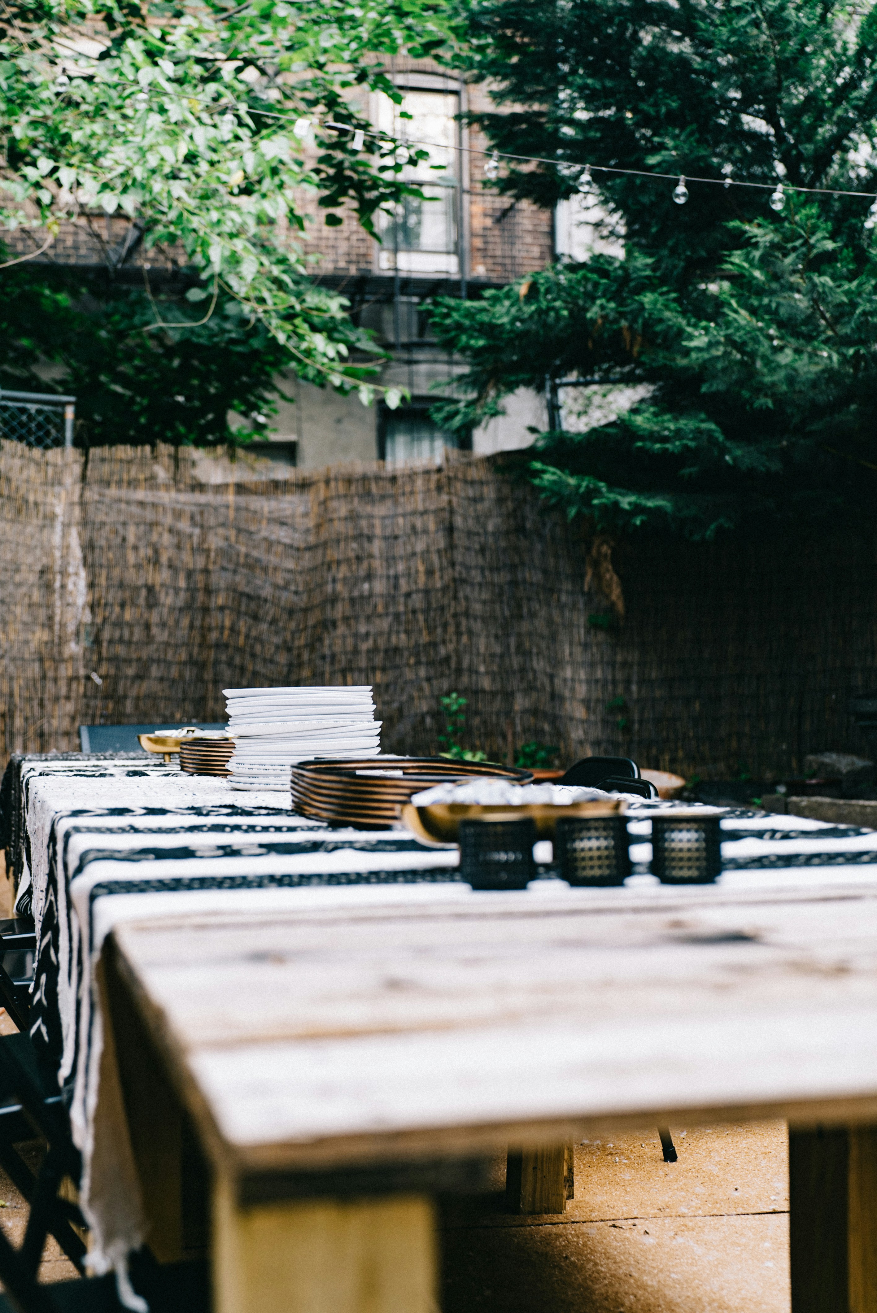 Outdoor dining setup featuring a long table adorned with plates and candles, surrounded by lush greenery and a rustic backdrop.