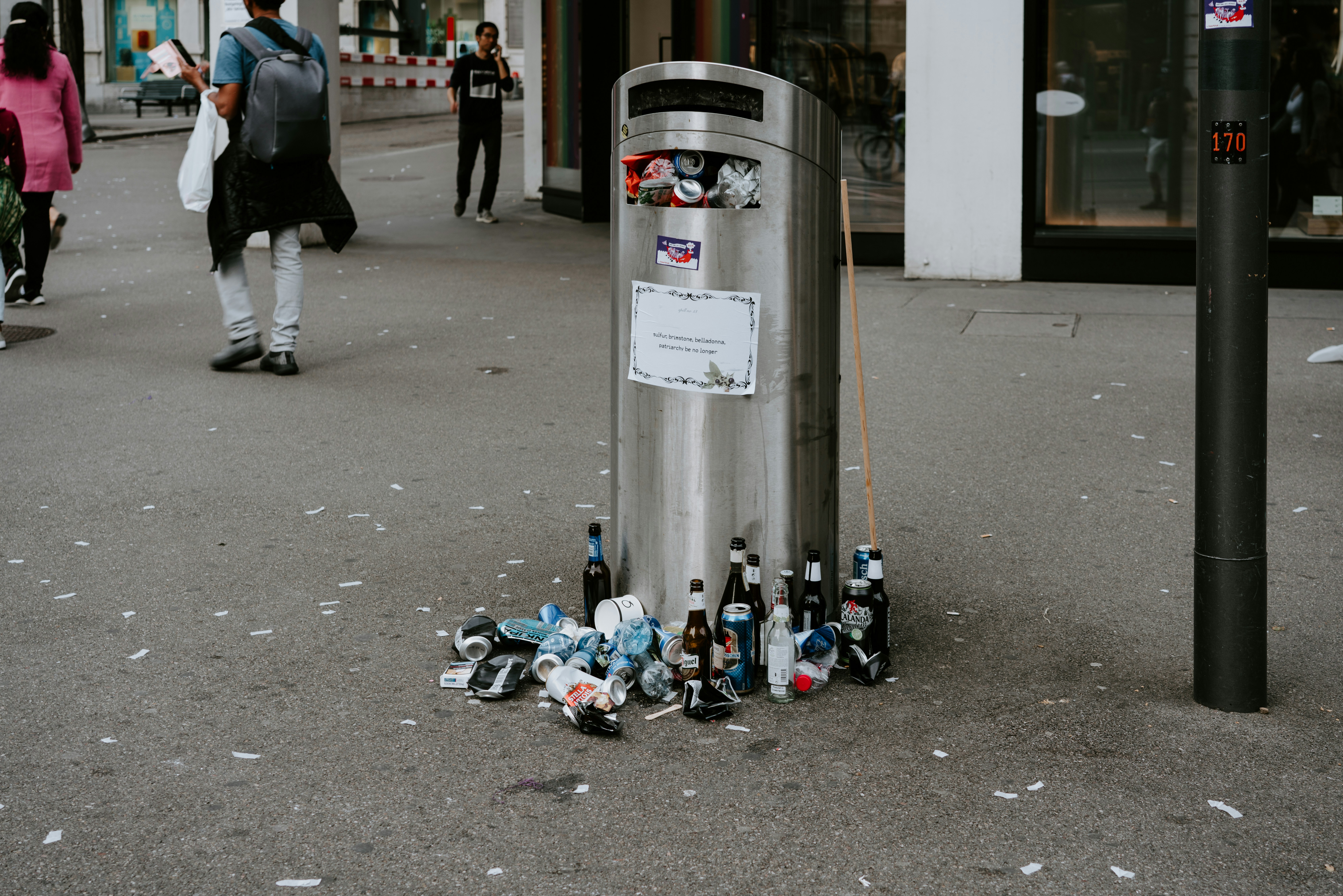Overflowing trash bin surrounded by discarded bottles and litter in a city street, highlighting urban waste management issues.