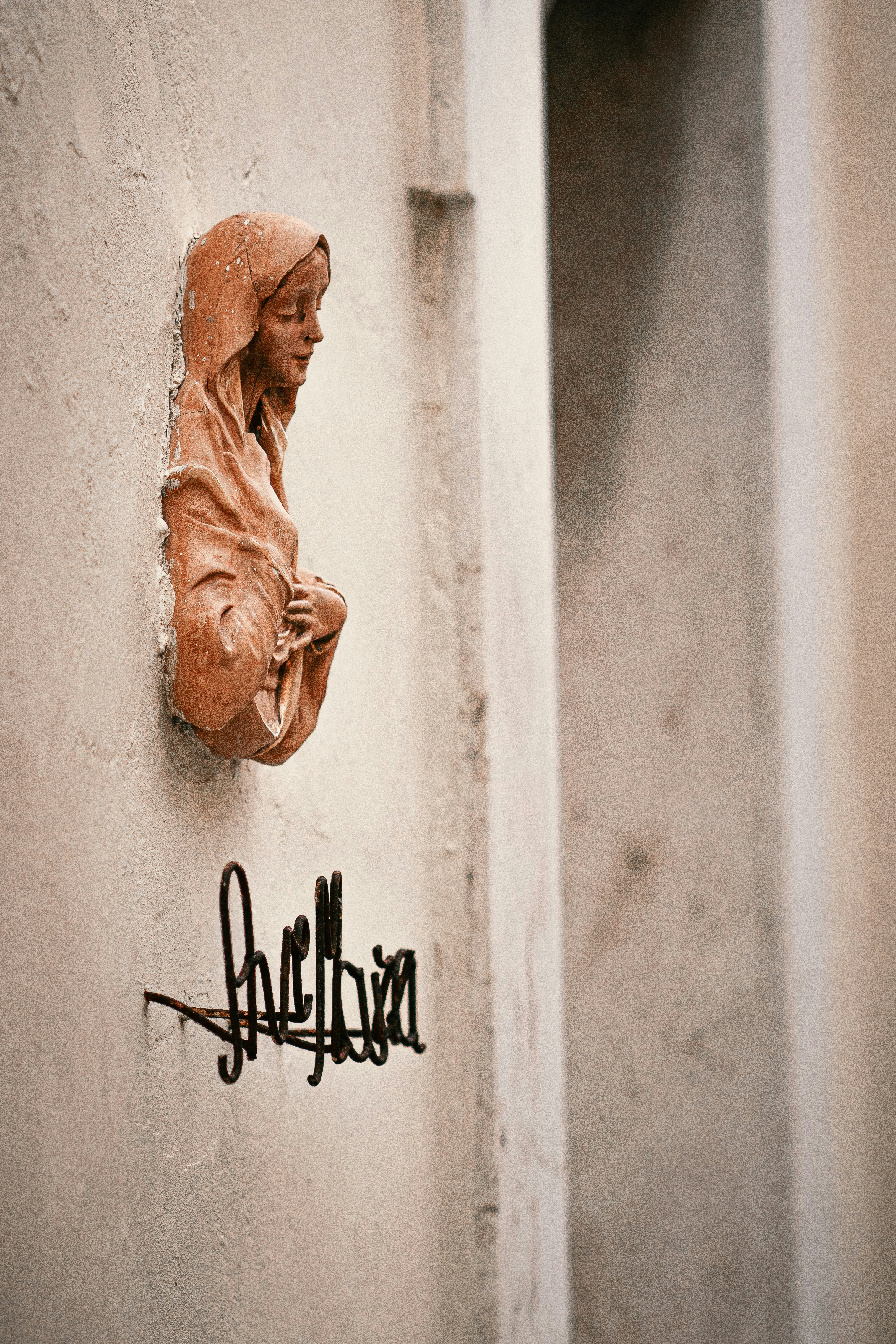 Terracotta sculpture of a woman in a hood, embedded in a textured wall, accompanied by a stylized signature. The piece blends artistry with urban surroundings.