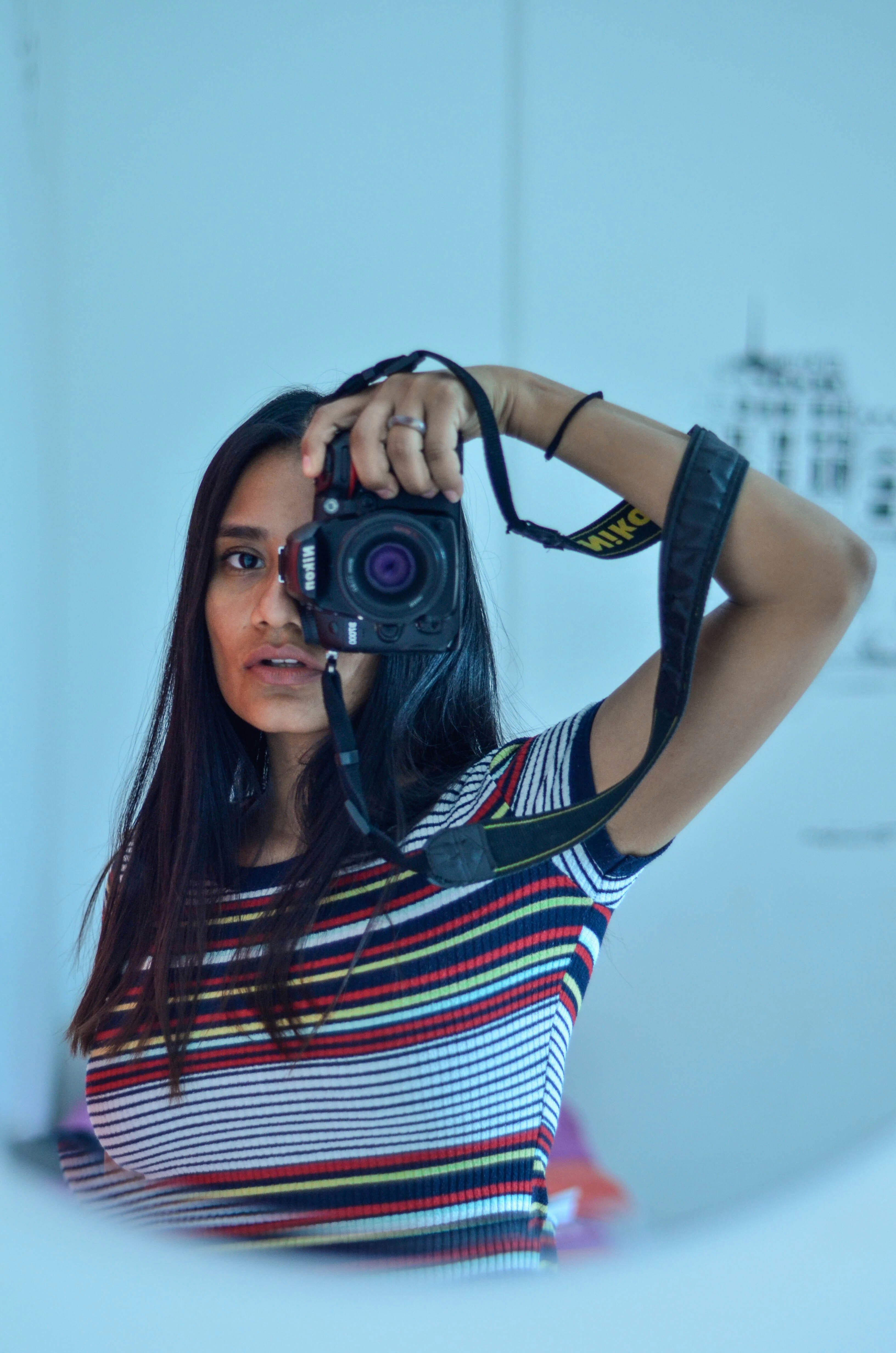 Photograph of a young woman holding a Nikon camera, taking a self-portrait in a circular mirror, wearing a multicolored striped shirt.